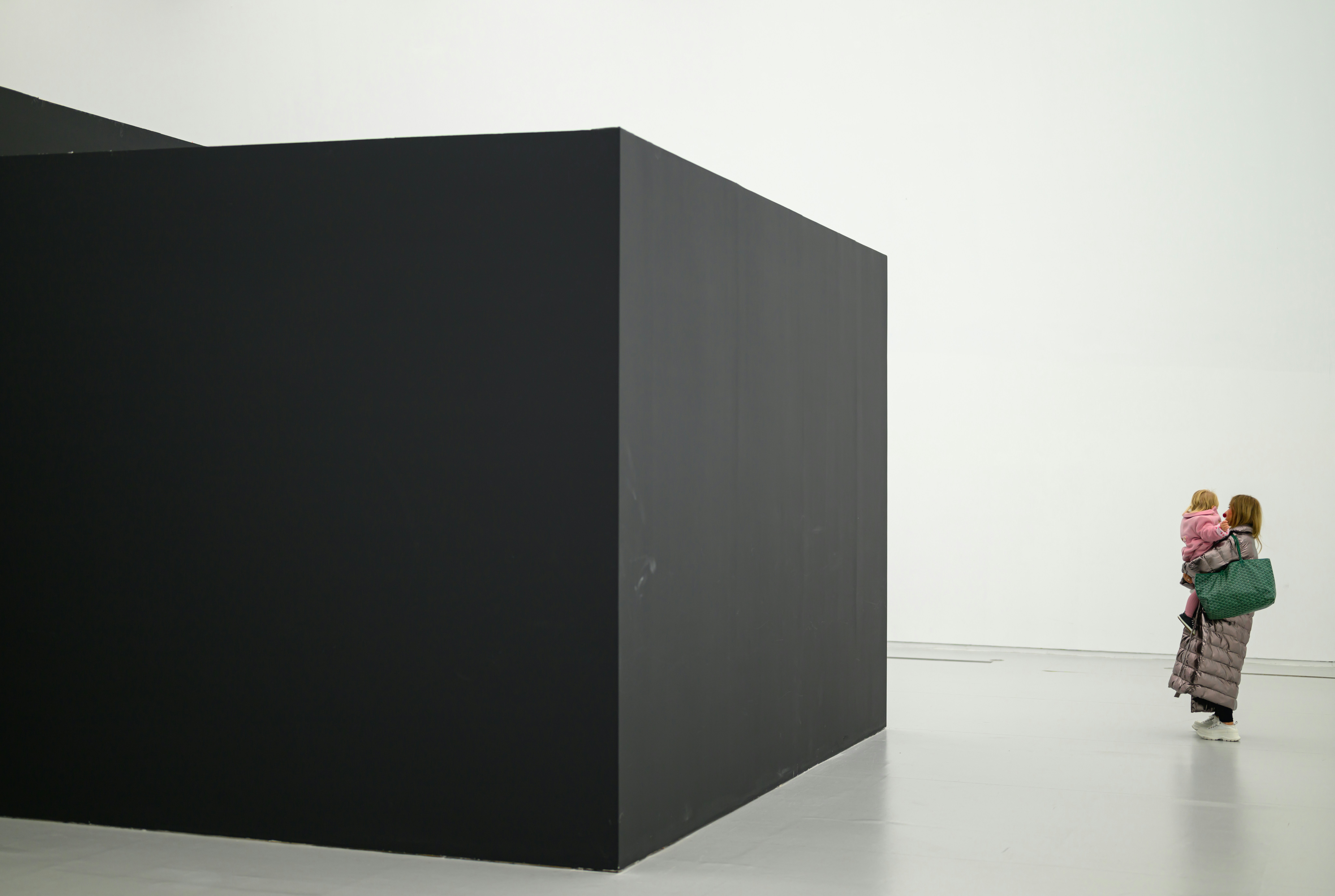 A couple stands near a large black rectangular sculpture in a white-walled gallery, their embrace softened by distance and pale lighting.