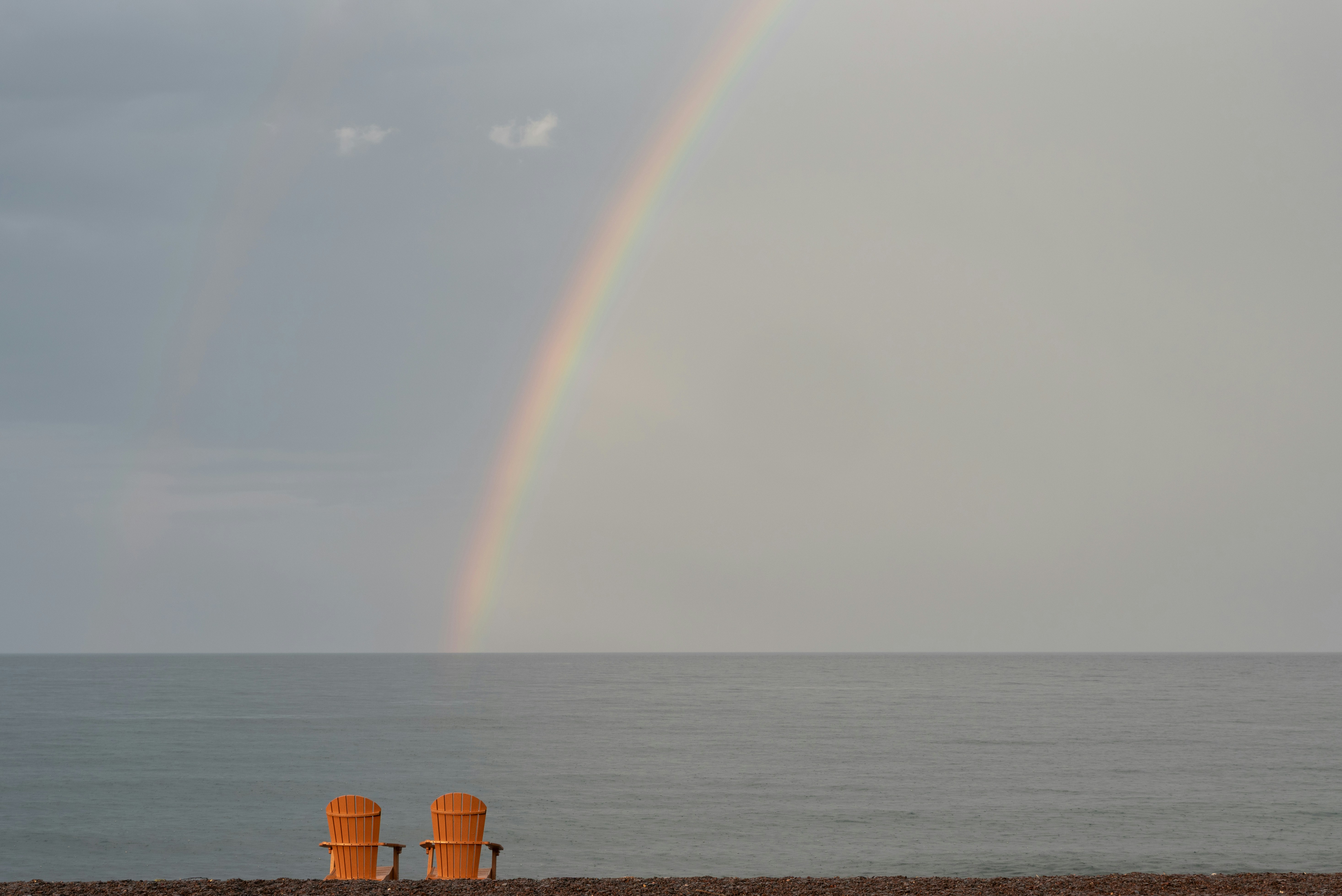Two bright orange Adirondack chairs sit side by side on a pebbly shoreline, facing a vast, glassy lake beneath a striking rainbow that arches boldly across a soft grey overcast sky. The simplicity of the composition — just chairs, water, sky, and rainbow — creates an immediately powerful sense of peace, companionship, and wonder. The warm pop of orange from the chairs against the cool grey and silver tones of the water and sky makes the image visually arresting. Perfect for travel, relaxation, companionship, hope, lakeside lifestyle, cottage living, summer getaway, or inspirational concepts.