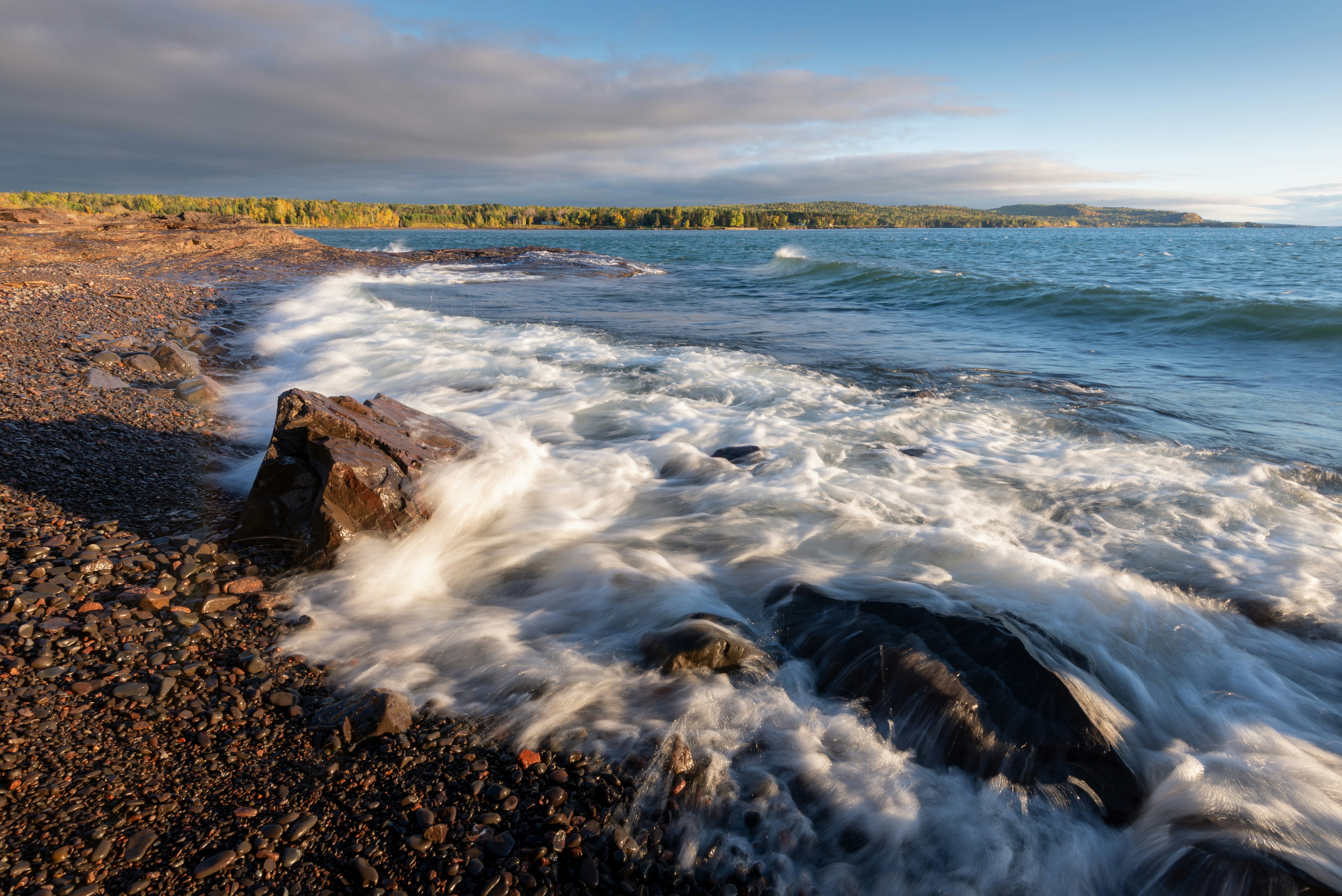 Dynamic waves surge and swirl around dark basalt rocks and a colorful pebble shoreline, captured with a slightly longer exposure that renders the rushing water as silky white foam and motion blur. The vivid teal-blue water of a large freshwater lake stretches to a distant forested shoreline glowing with early autumn color — golden yellows and deep greens beneath a partly cloudy blue sky. The wide-angle perspective pulls the viewer right into the action at the water's edge, creating a powerful sense of immersion and energy. Perfect for Great Lakes, wilderness, adventure, nature, travel, autumn, water power, or dramatic landscape concepts.
