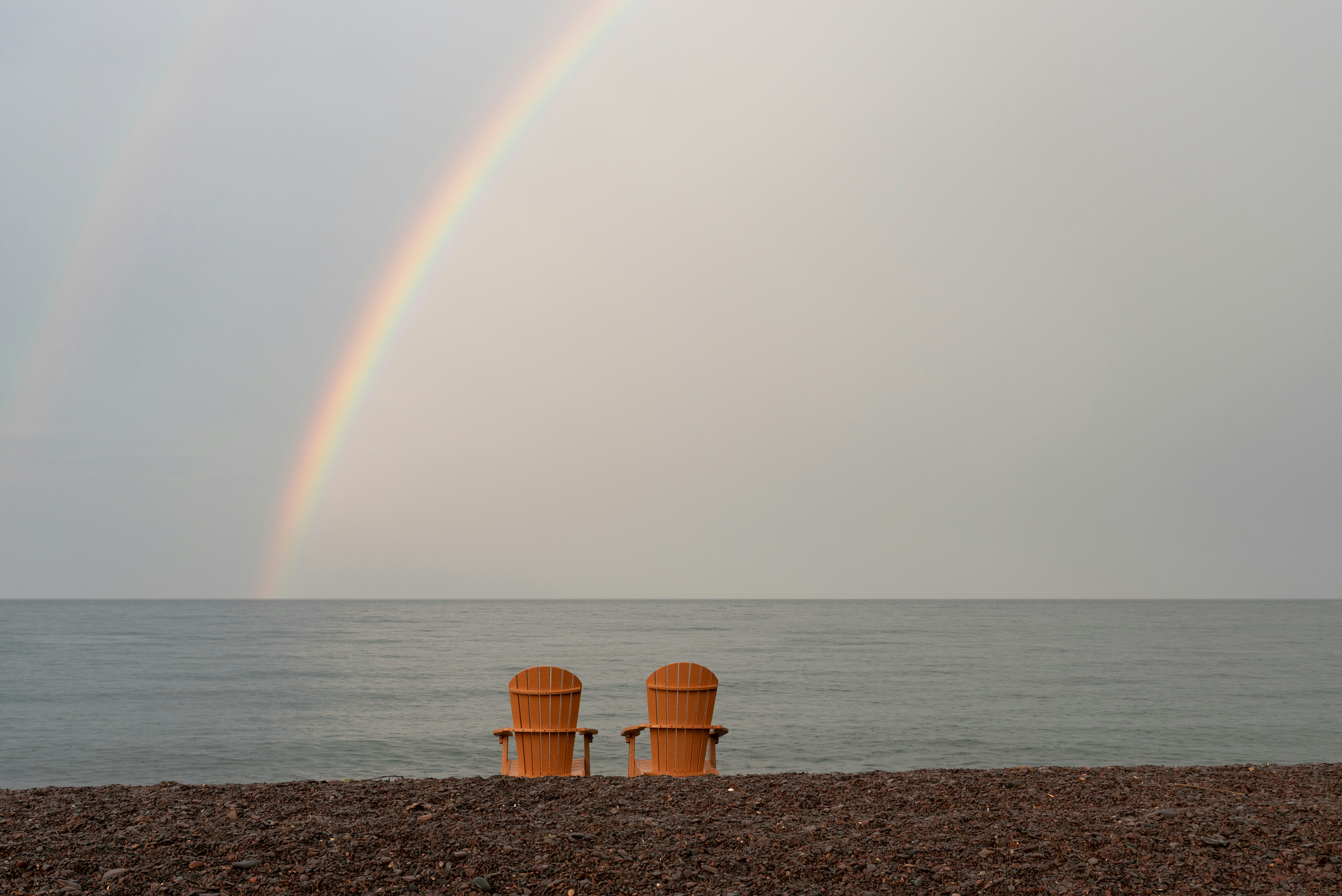 Two Adirondack Chairs Beneath a Double Rainbow Over a Quiet Sea