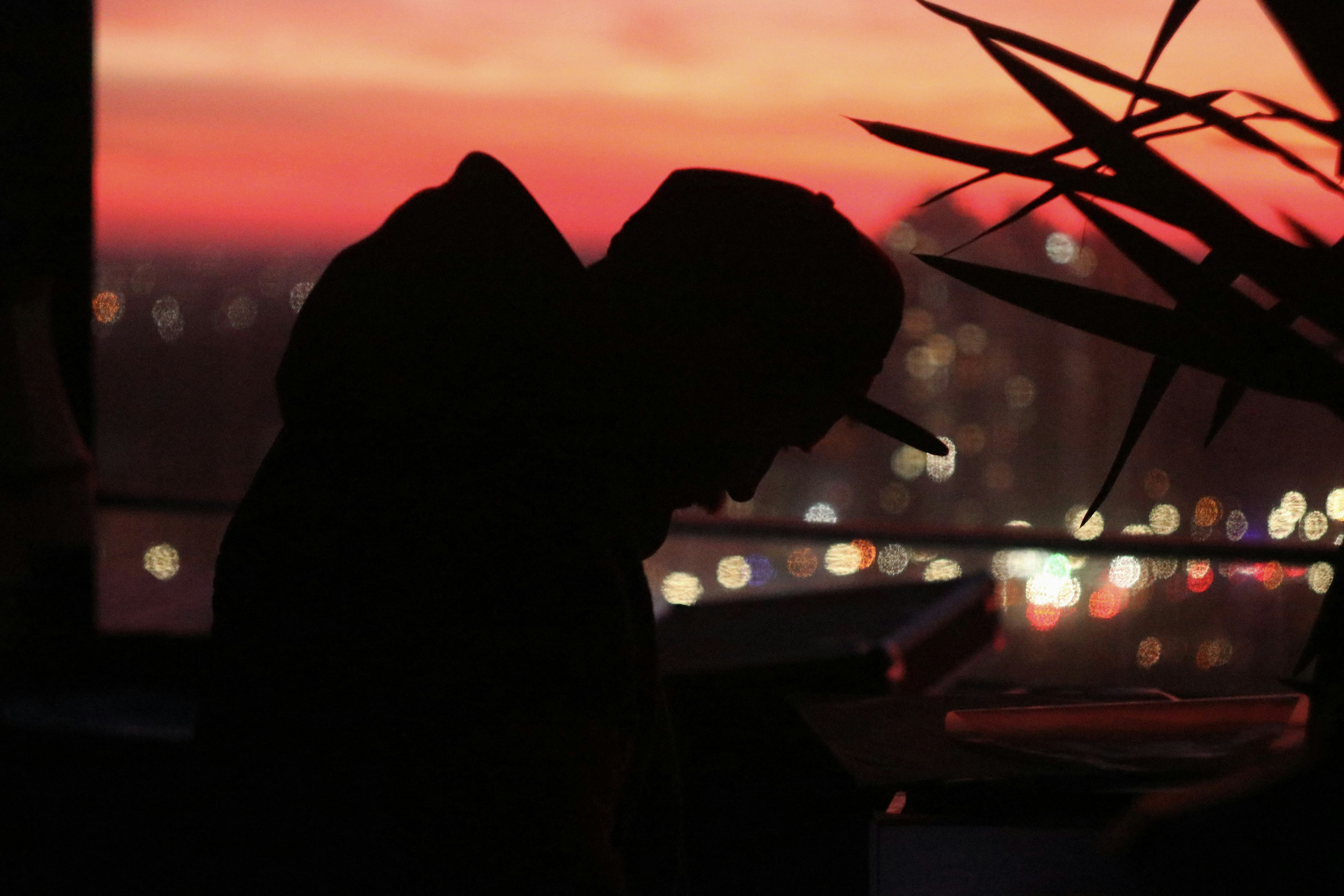 A moody silhouette of a person wearing a cap against a vibrant, fiery sunset sky. The background features blurred city lights creating beautiful bokeh, capturing a cinematic, contemplative urban atmosphere.
