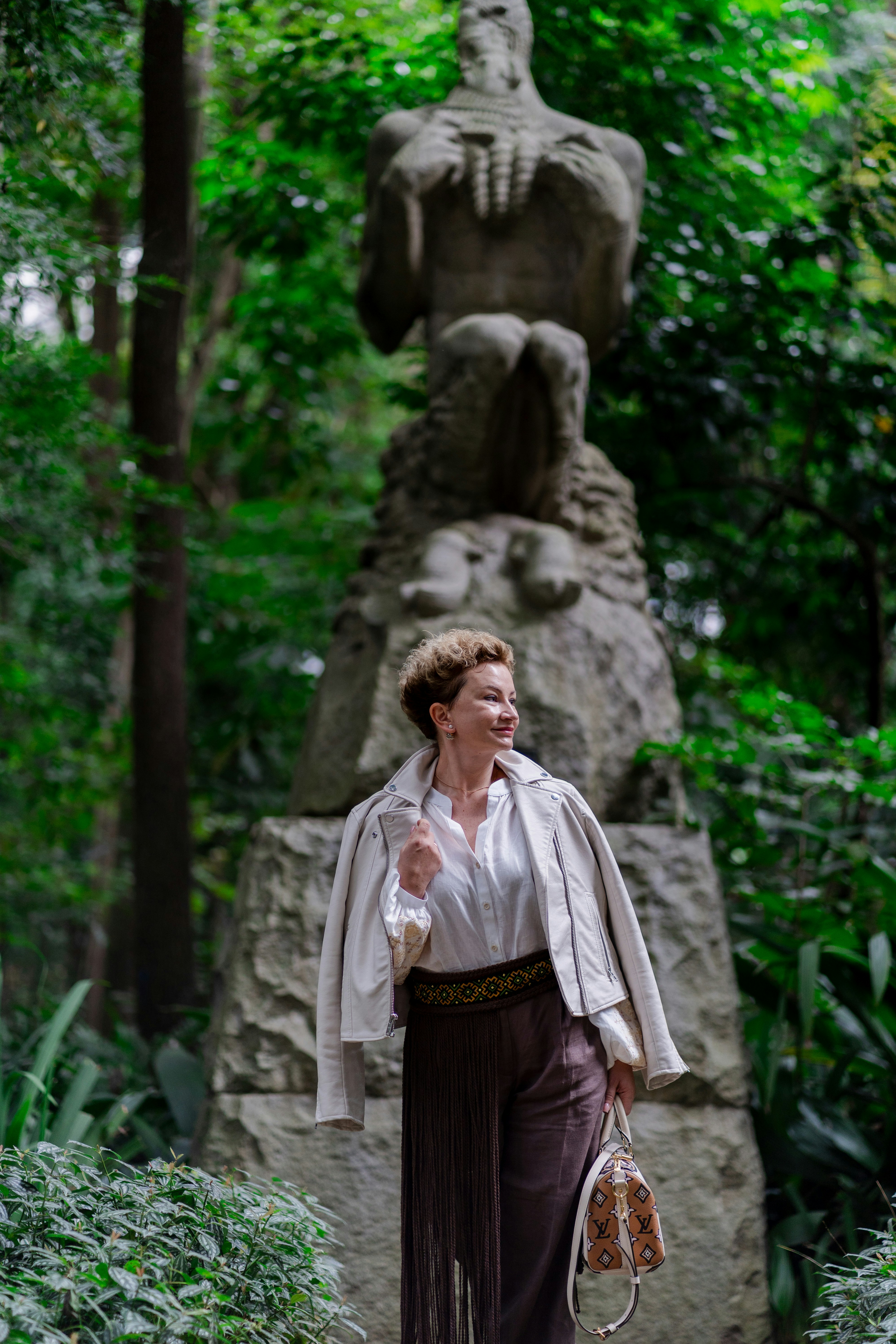 Woman in colonial blouse and fringe belt walks past a weathered statue in a lush garden