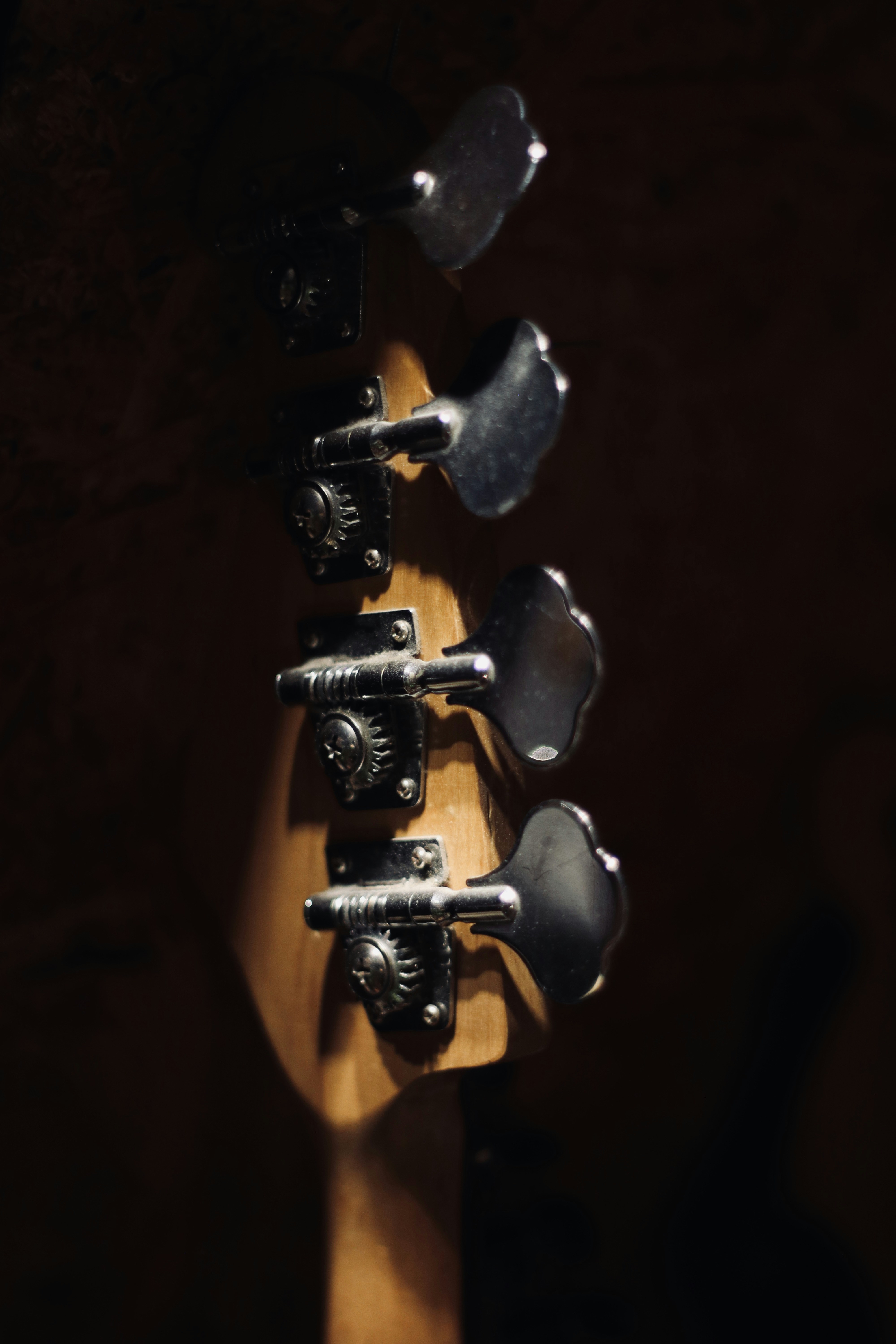 A close-up, high-contrast shot of the headstock and chrome tuning pegs on a wooden bass guitar.