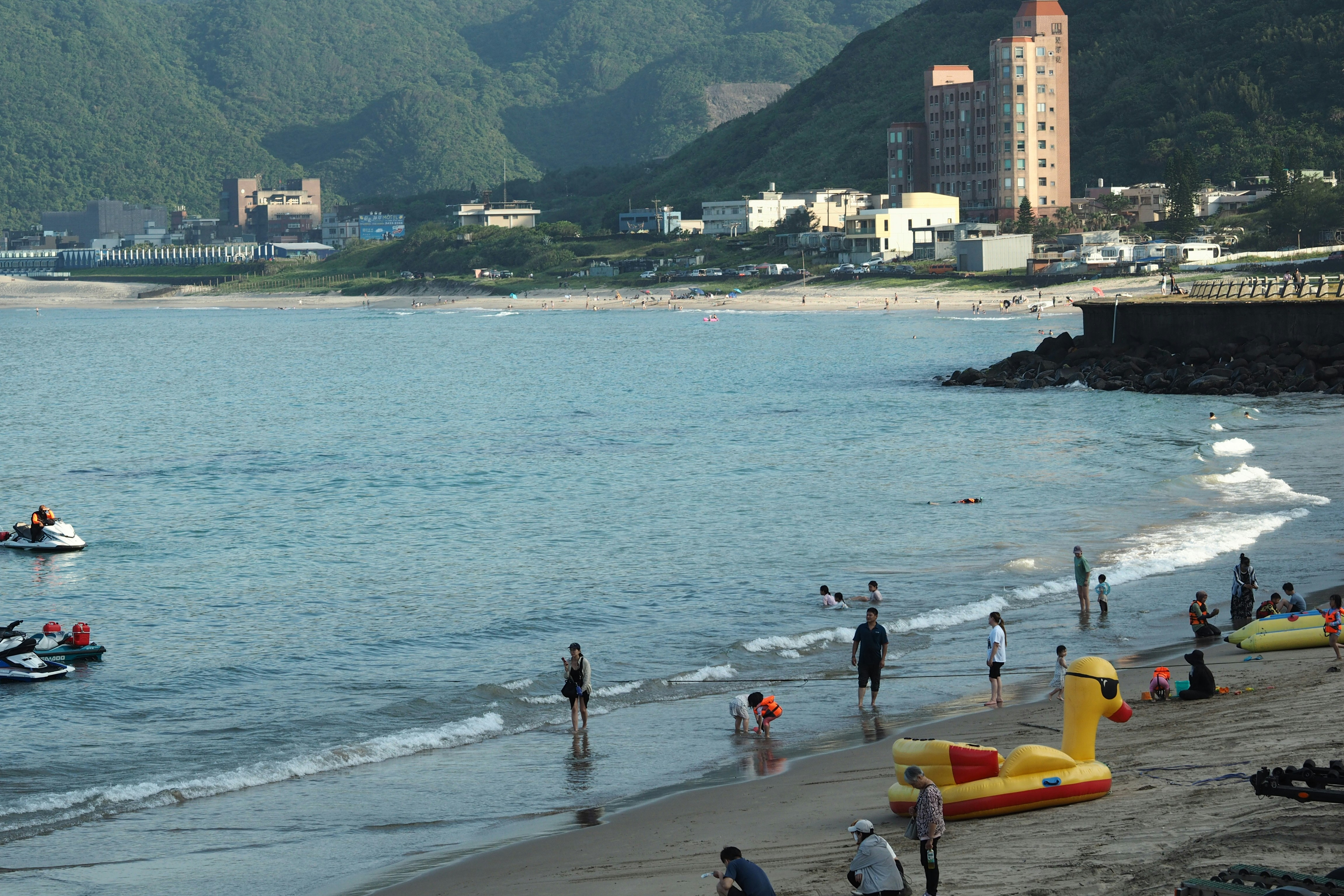 Sunlit shoreline with swimmers and a buoyed inflatable near a quiet urban coast
