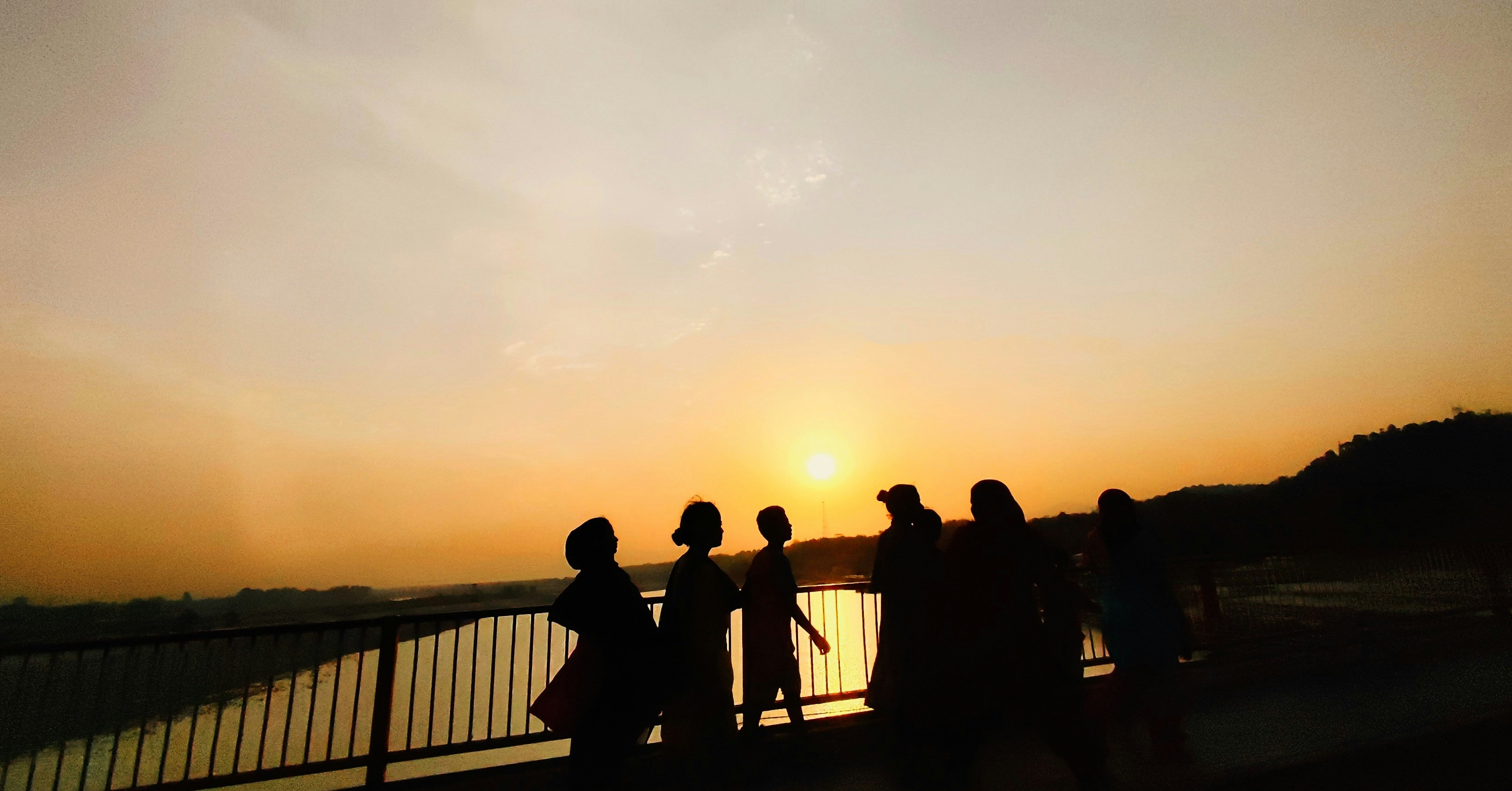 A group of people walk across a bridge as the morning sun rises behind them, Their silhouettes contrast against the glowing sky, creating a candid and atmospheric scene that captures the beauty of everyday moments.