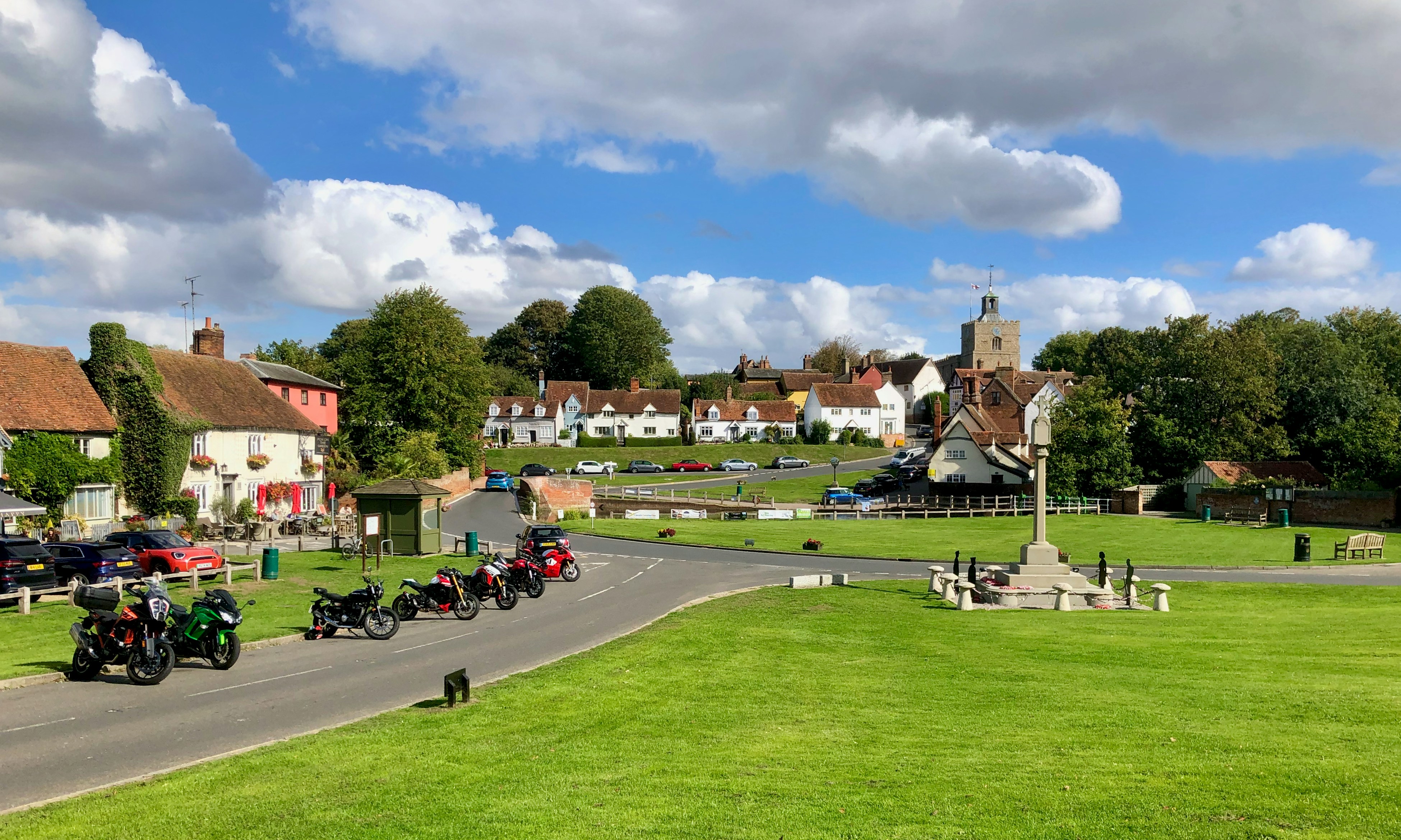 Bright town square with a central monument, surrounding cottages and parked motorcycles on a curved road; expansive green lawns and a blue sky with fluffy clouds.