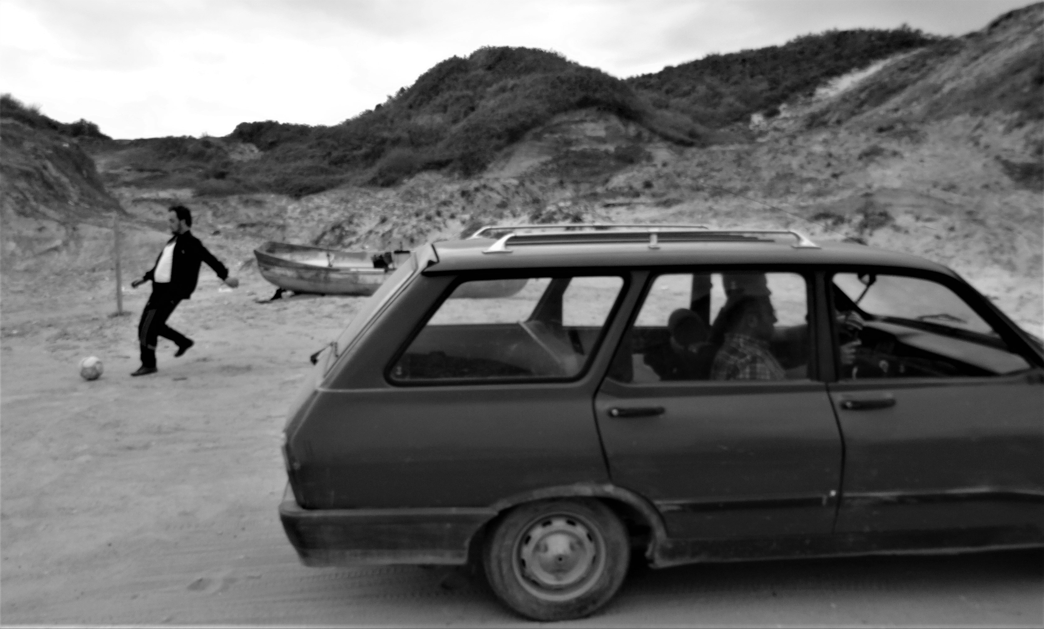 Dusty Beach Run: A Booted Figure and a Parked Wagon in Quiet Monochrome