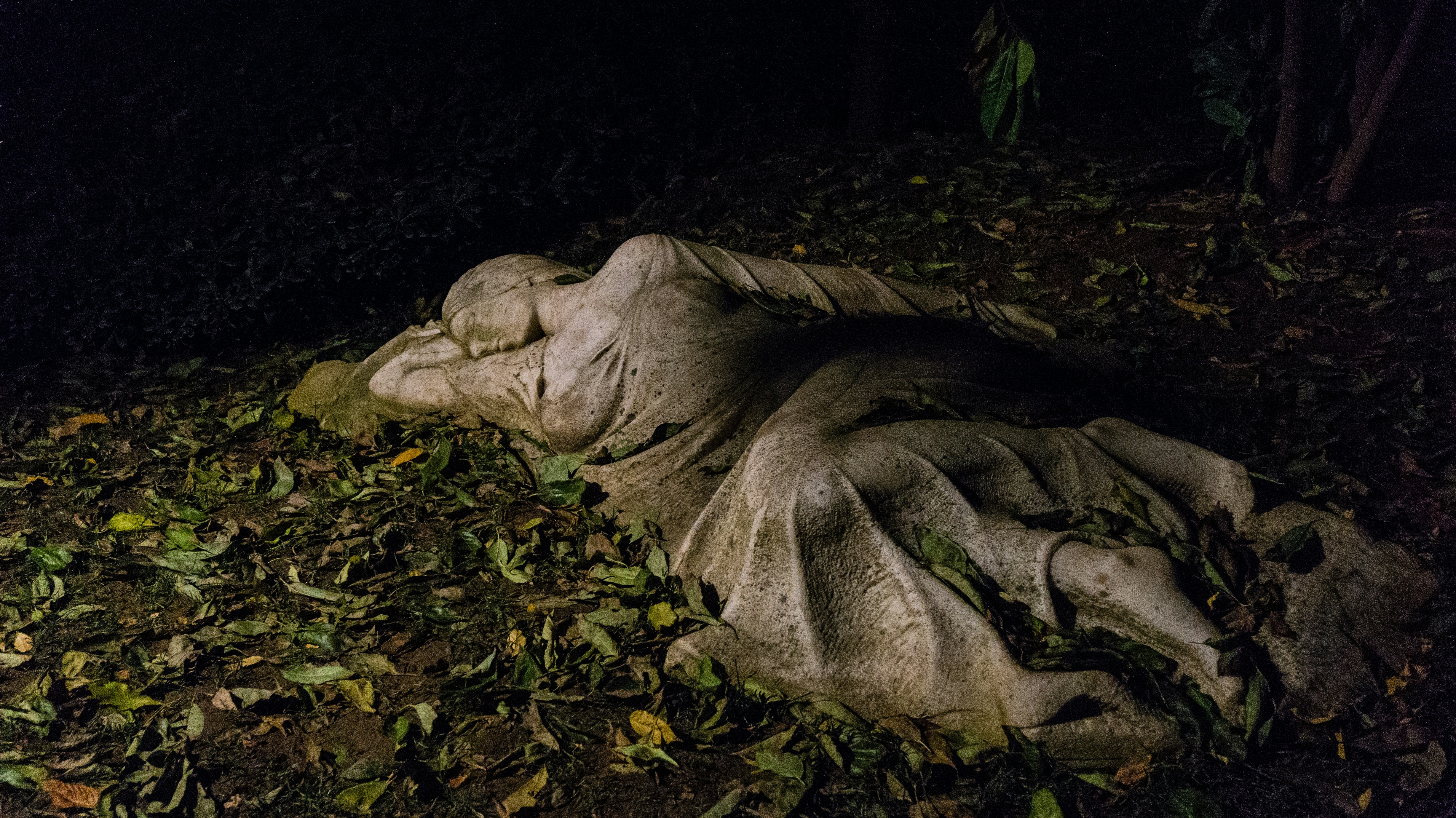 A weathered stone statue lies on leaf-covered ground in darkness.