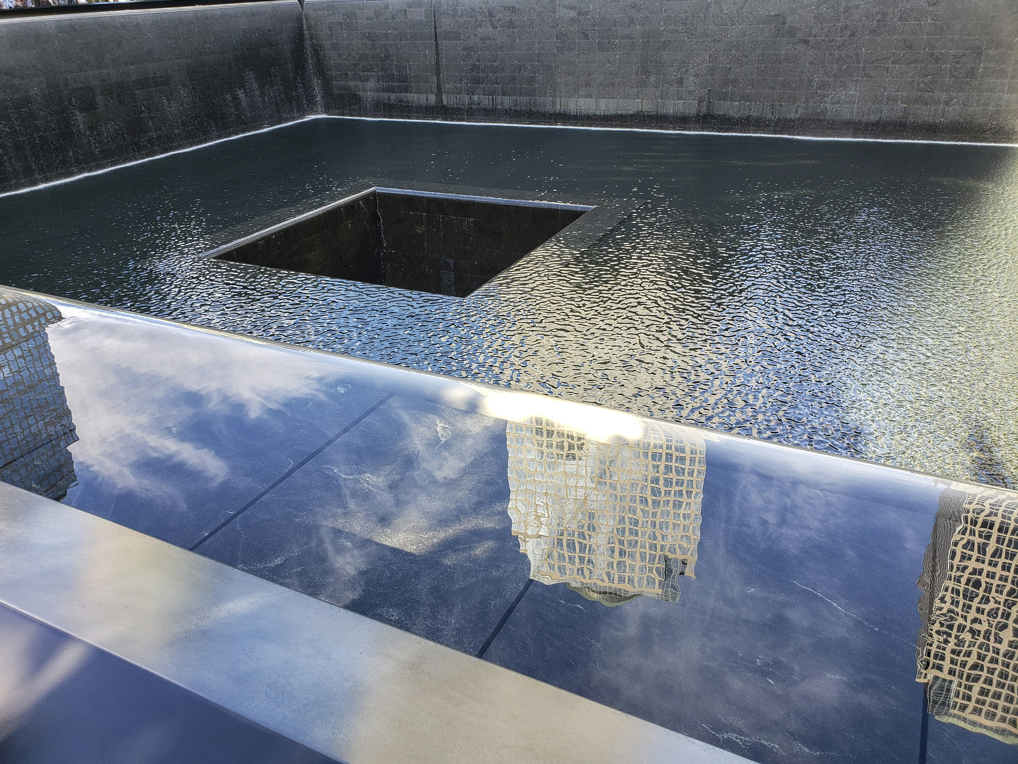 Reflective pool with a square central opening, surrounding dark walls, and the reflected cityscape on its glossy surface.