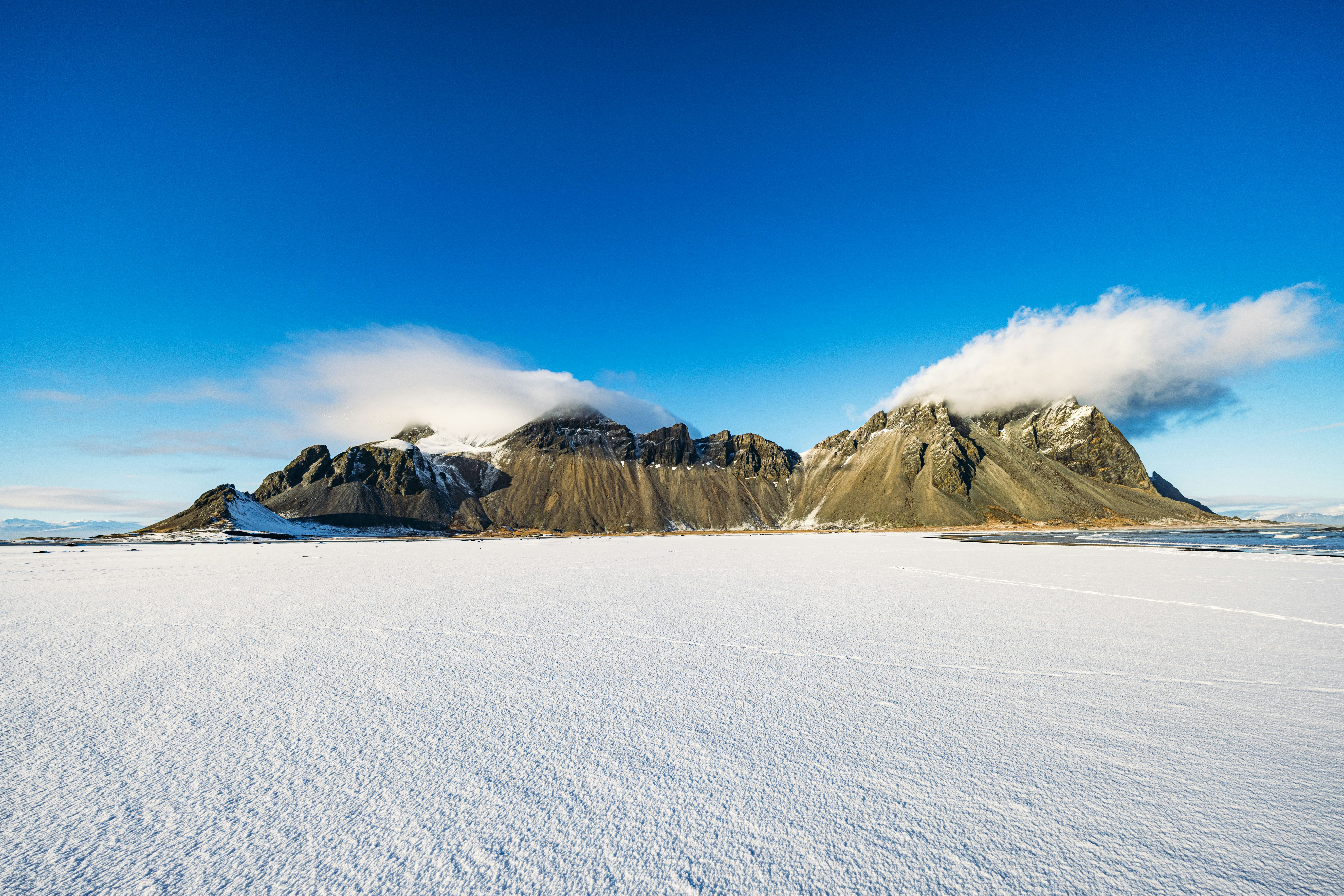 Vast snowfield before rugged mountains with clouds under blue sky.