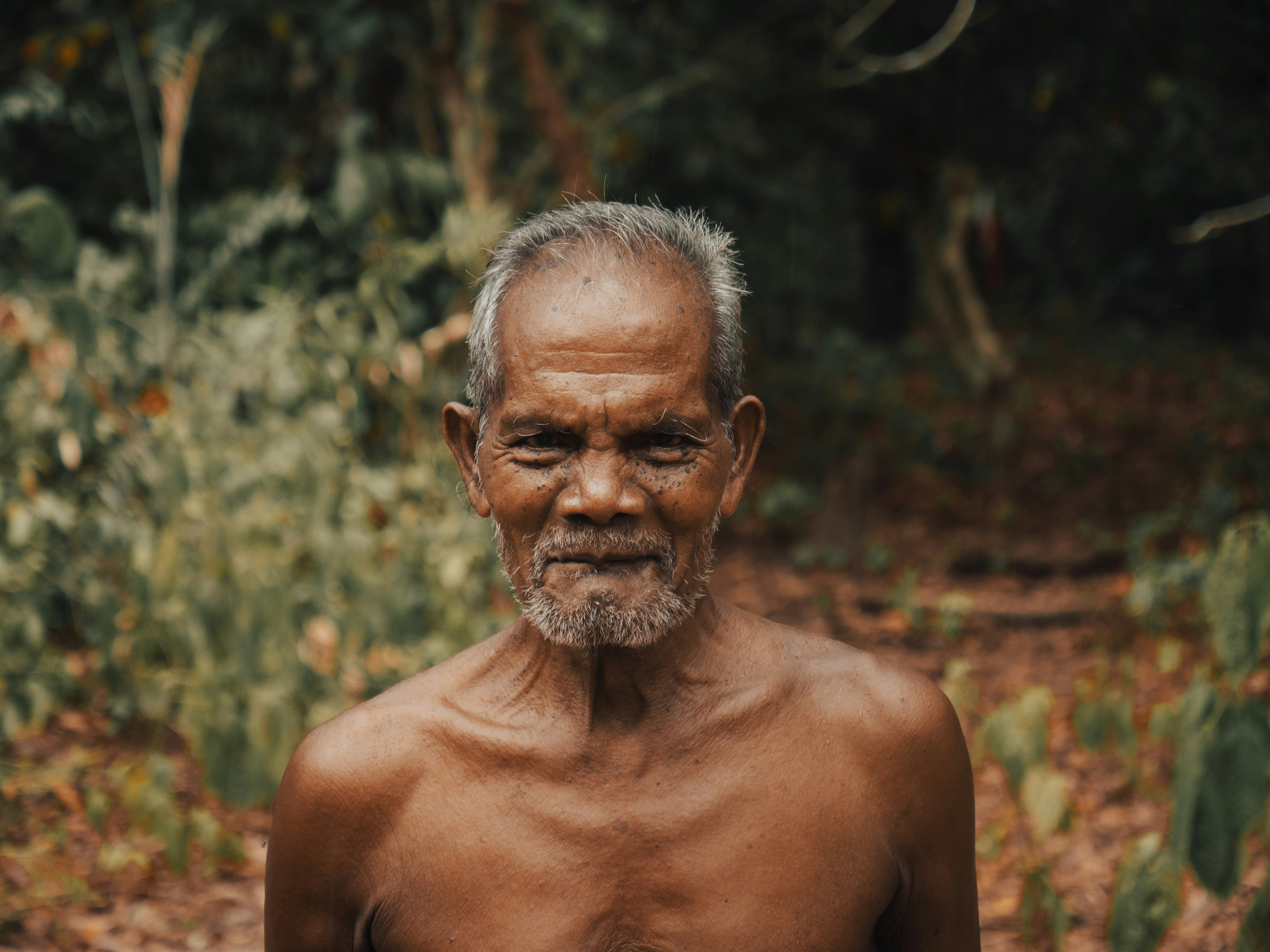 Bare-chested elderly man with a beard in a lush forest.