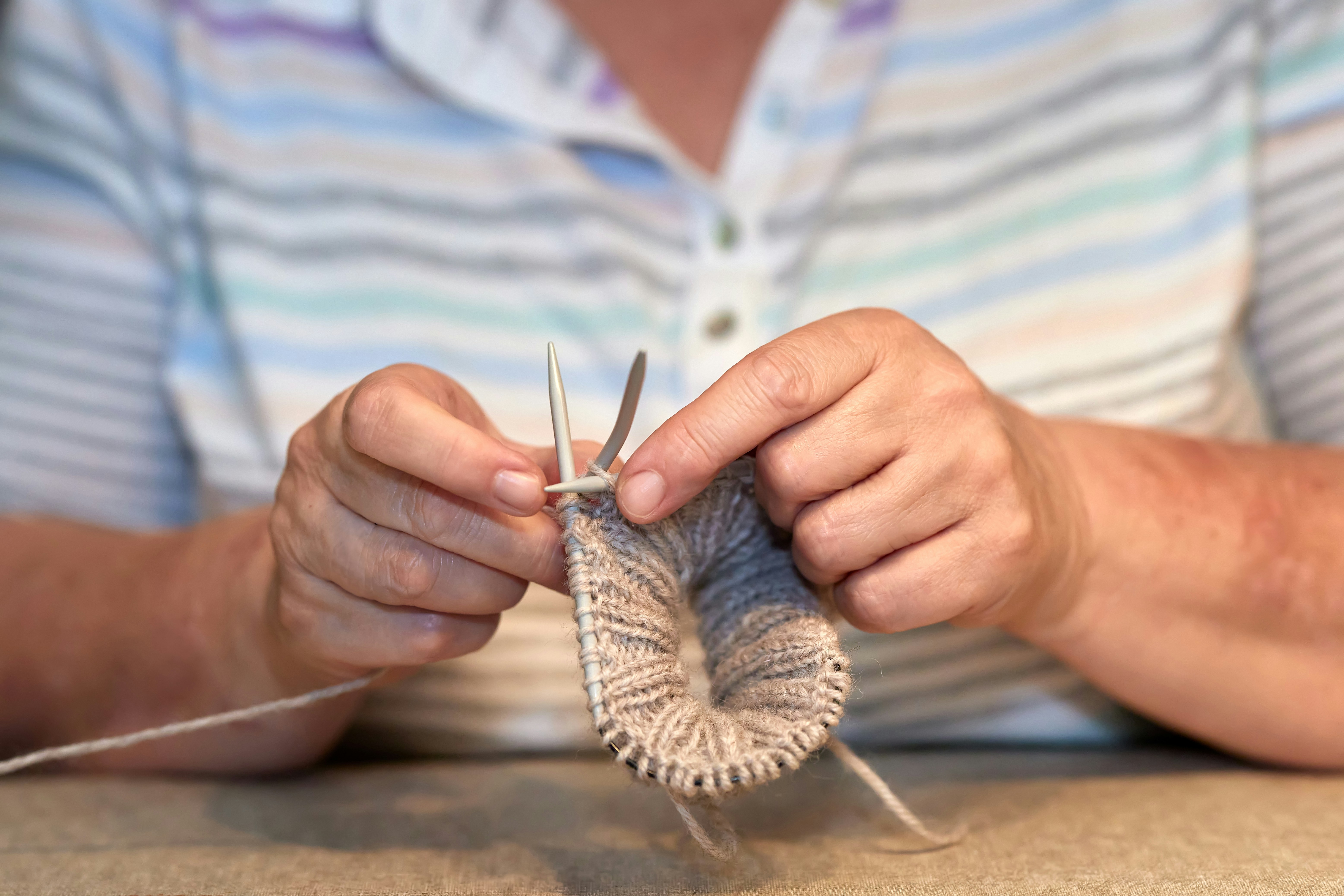 Close-up of hands knitting with light brown yarn and circular needles, focused on the creative process of making a handmade item. A relaxing and timeless hobby.