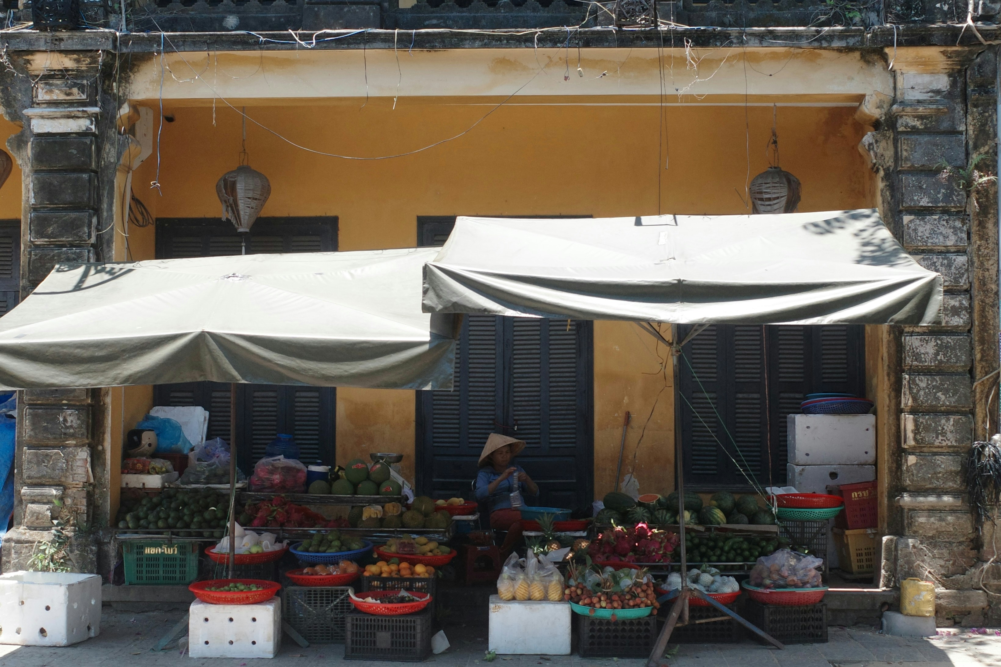 Vendor under Awnings: Market Stall with Fruits and Vegetables in a Sunlit Street