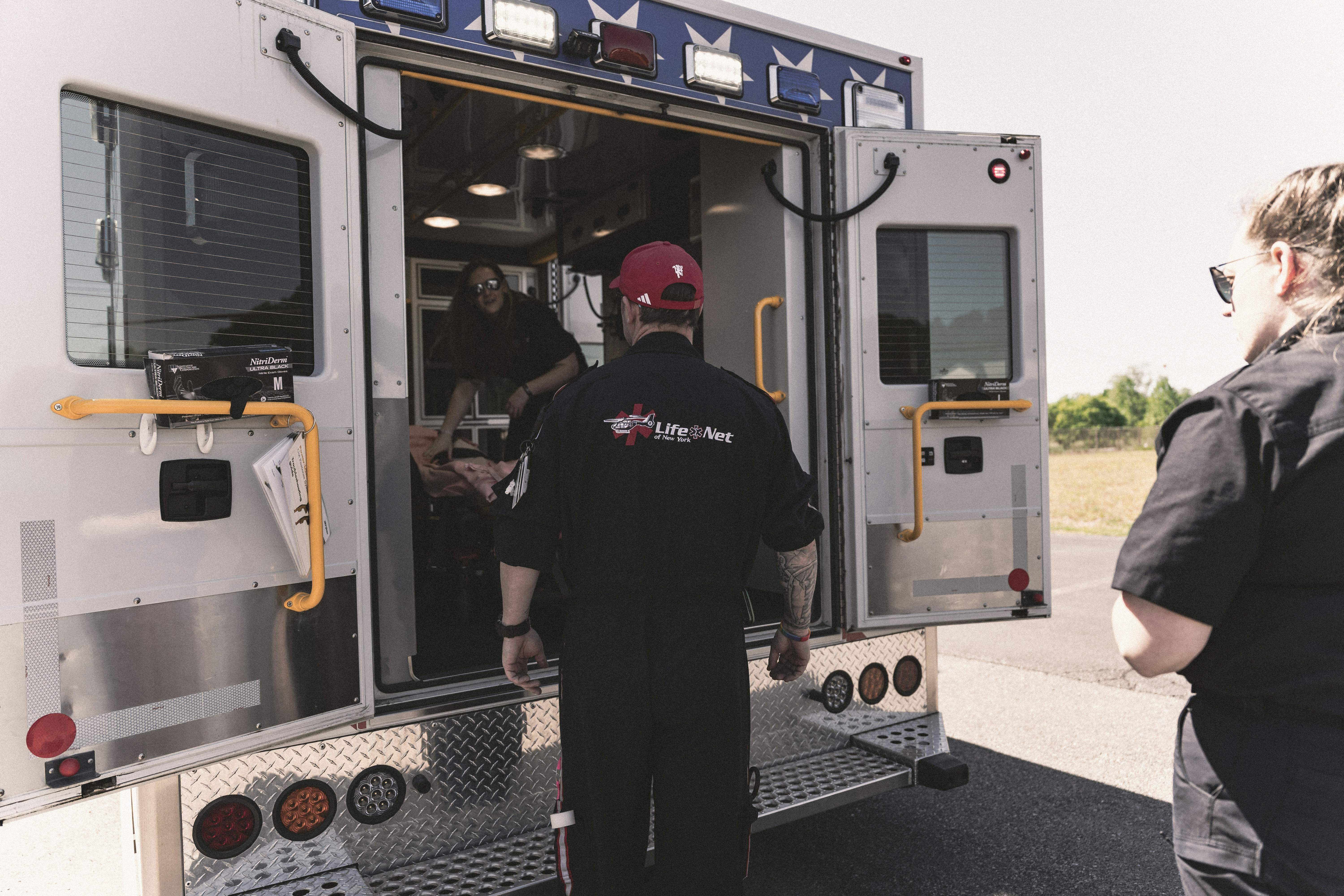Paramedics wheel a patient from an ambulance door, with a responder in red cap guiding the scene; a teammate stands by as the exterior light spills onto the tarmac. This is a documentary-style medical response scene.
