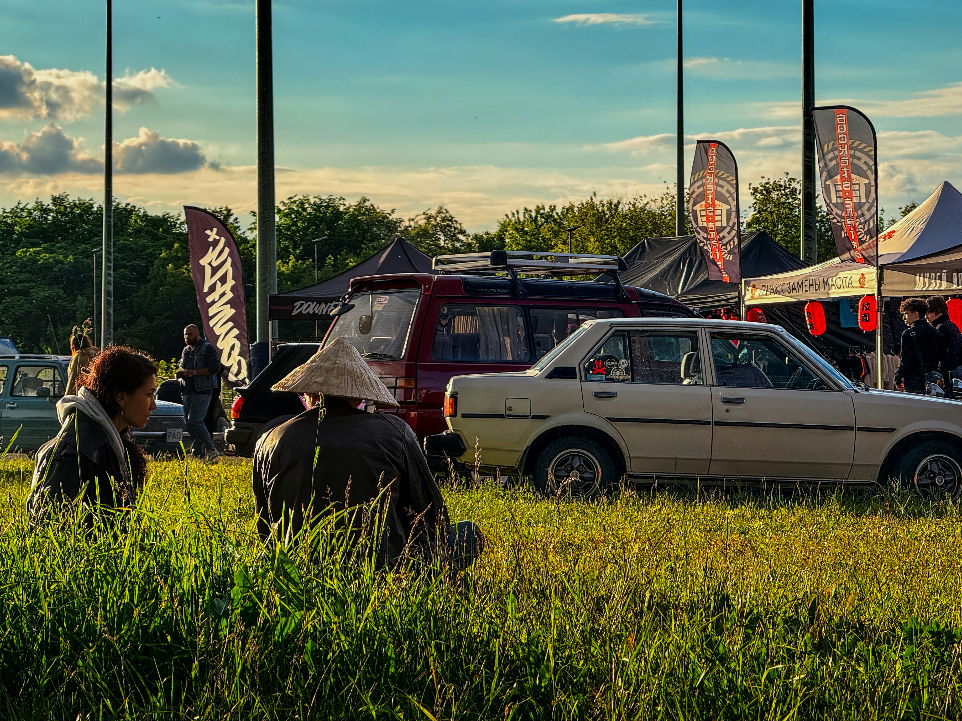 People sit in tall grass at an outdoor car show.