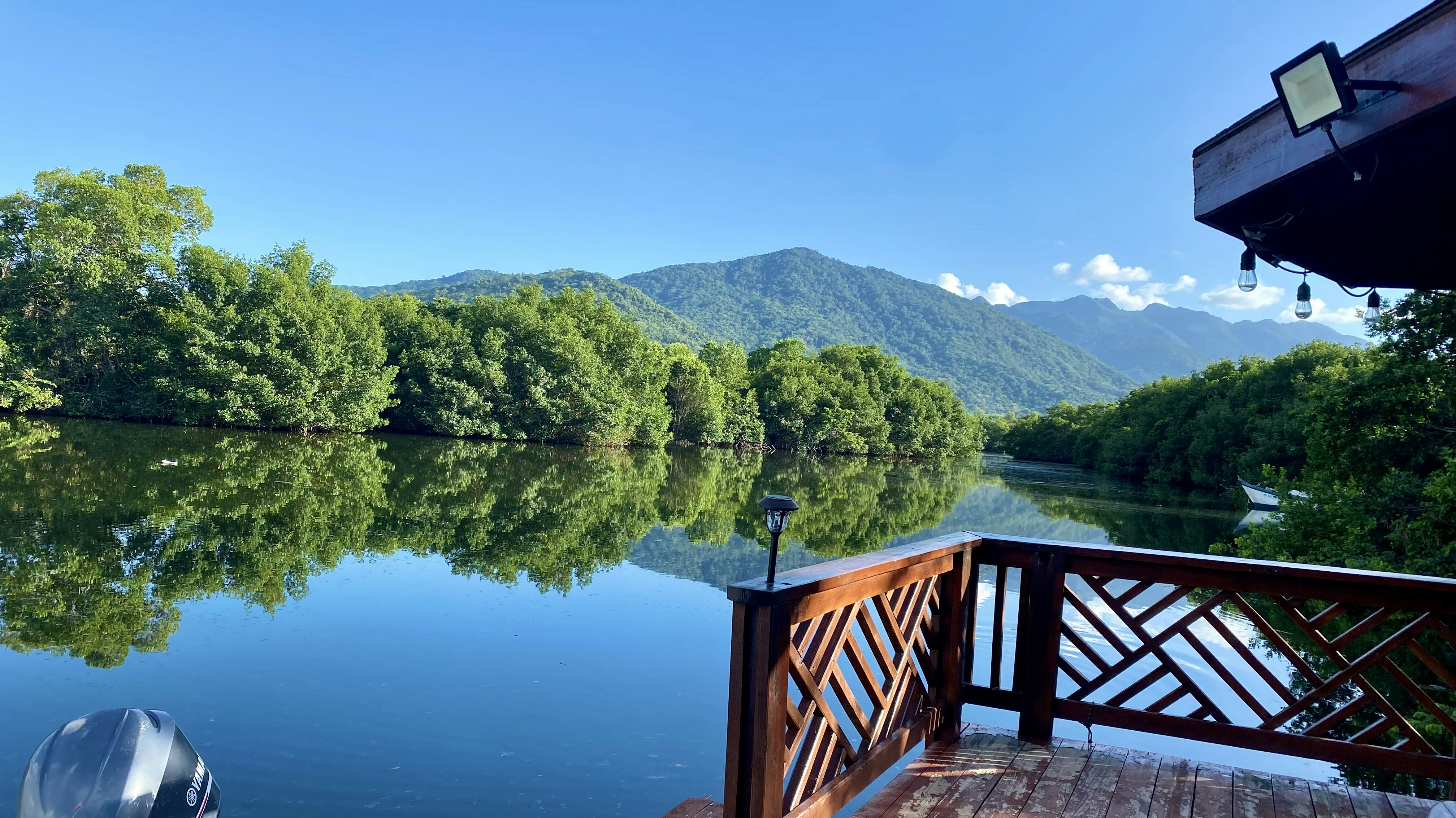 Calm river stretches into the distance with a lush tree line, a wooden deck railing in the foreground, and a distant mountain range under a bright sky.