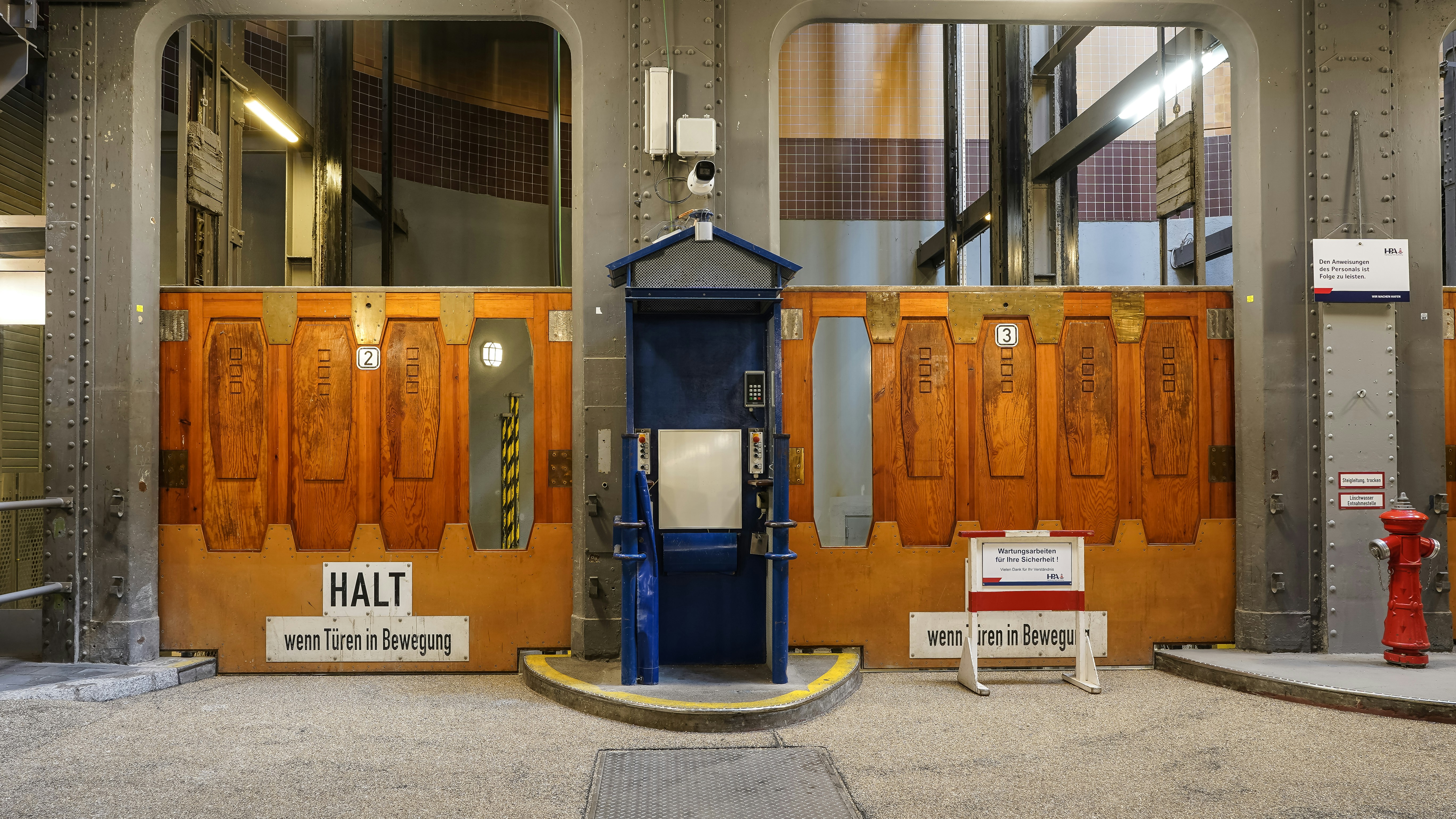Blue security booth centered between wooden doors in an industrial transit hall