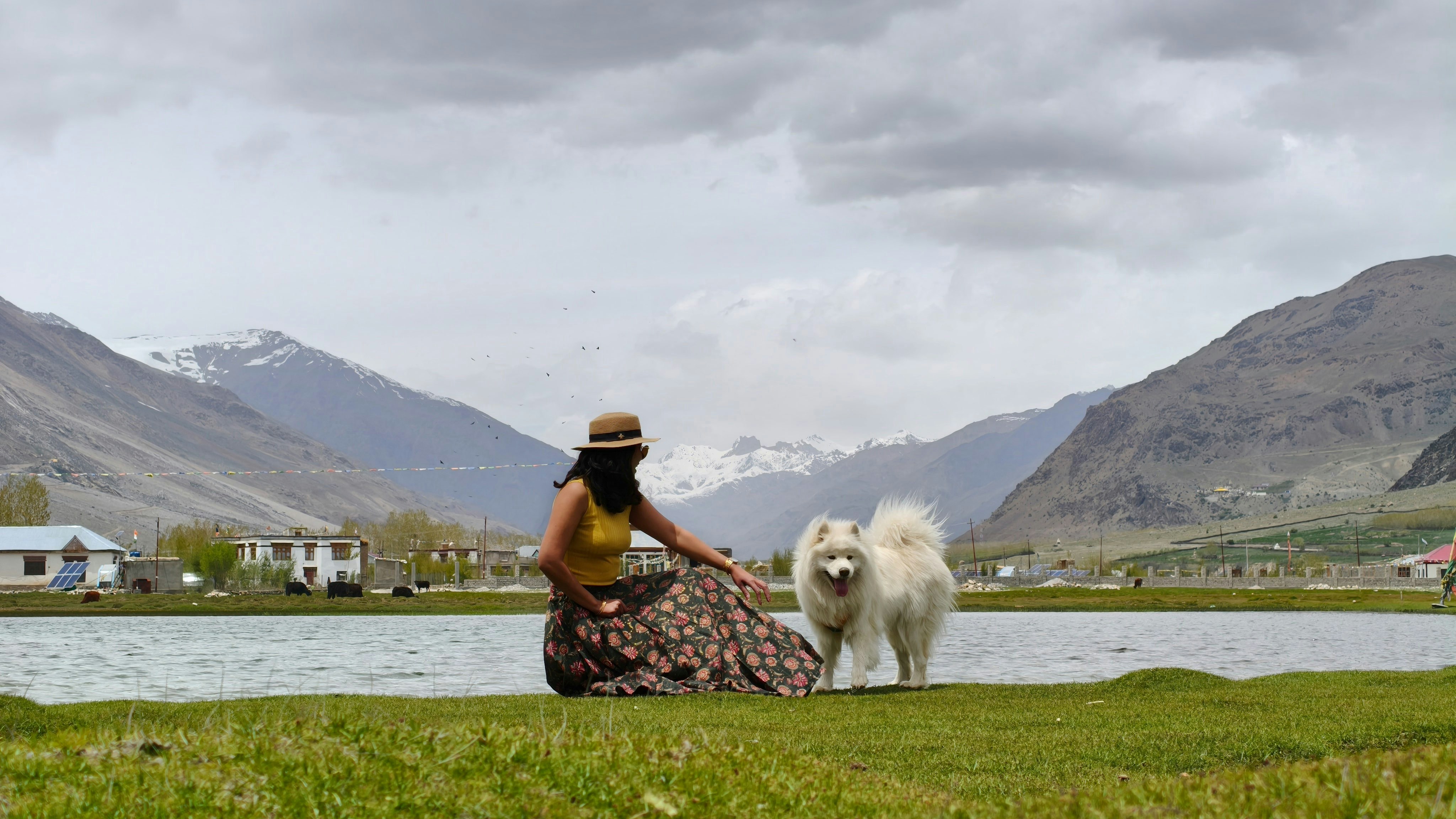 Woman in a floral skirt and hat with fluffy white dog near a lakeside valley