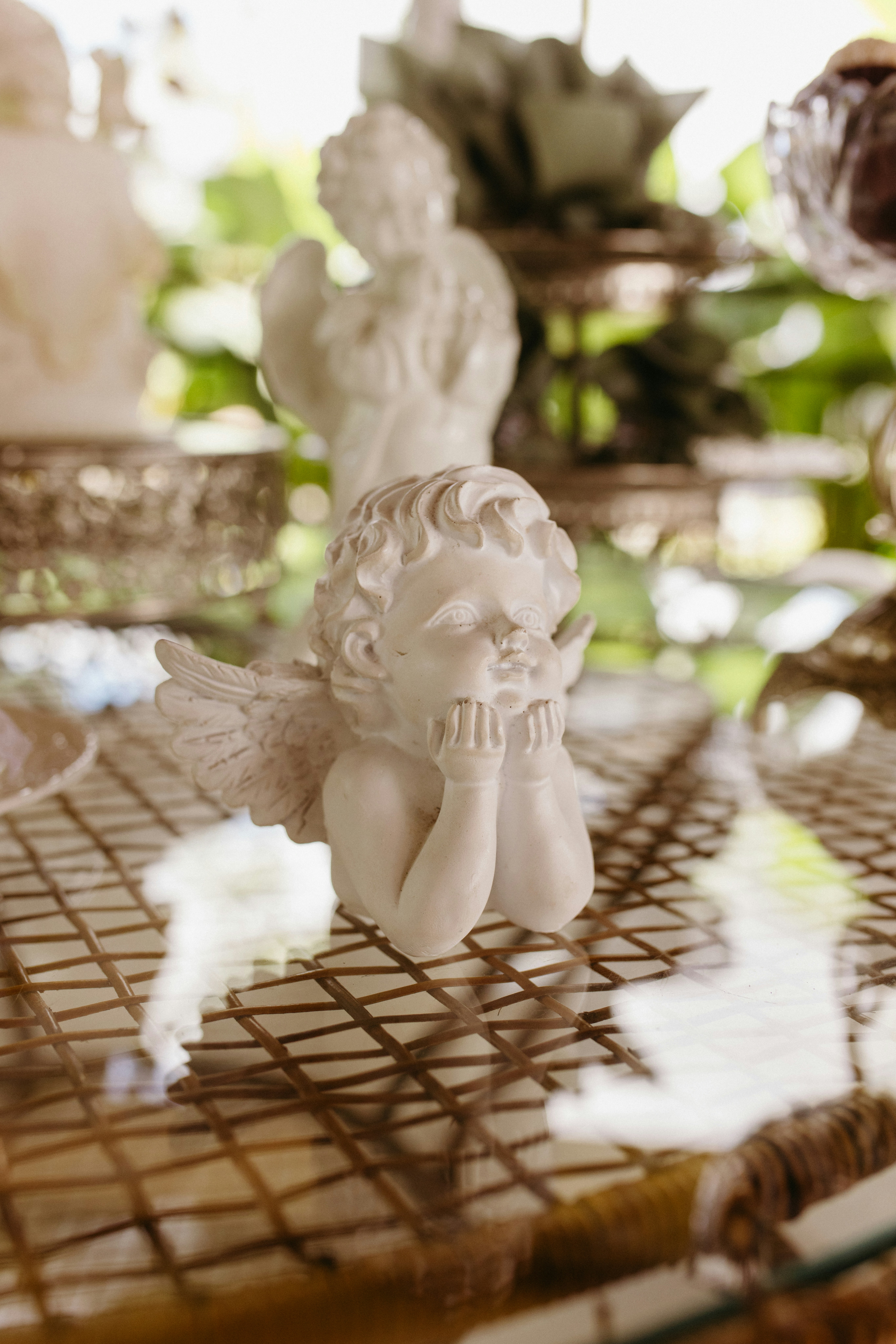 Small angel statue with clasped hands sits on a woven glass table, softly lit with blurred background decor including other statues and greenery.