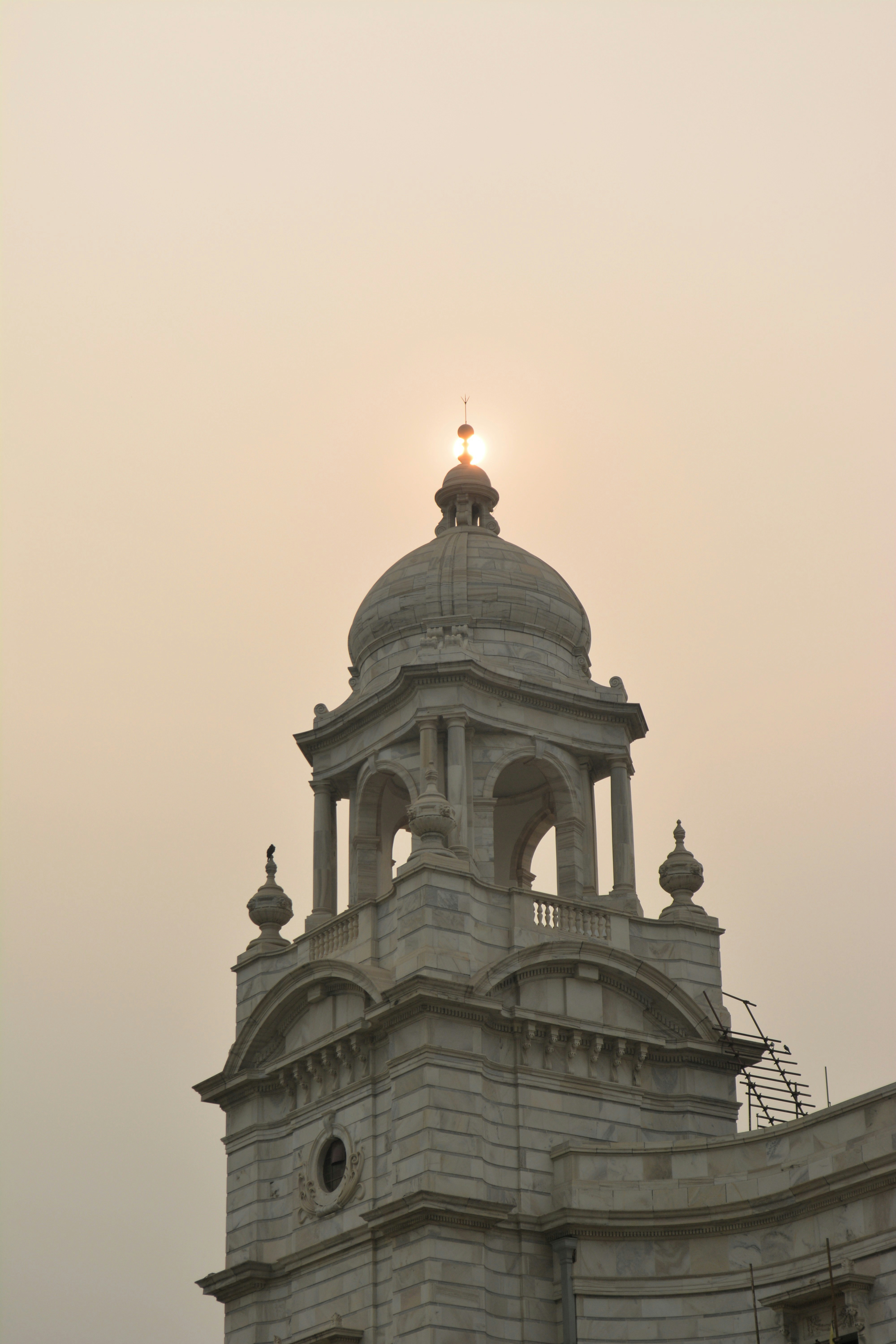 A striking architectural study of the iconic Victoria Memorial in Kolkata, West Bengal. The shot focuses on an ornate white Makrana marble dome and tower, showcasing its intricate British and Mughal-inspired Indo-Saracenic architecture. The hazy, golden sun is positioned precisely behind the topmost finial, creating a soft, ethereal backlight against a pale, warm morning sky. This minimalist composition highlights the grand detail, symmetry, and historical elegance of one of India's most famous monuments during a quiet sunrise.