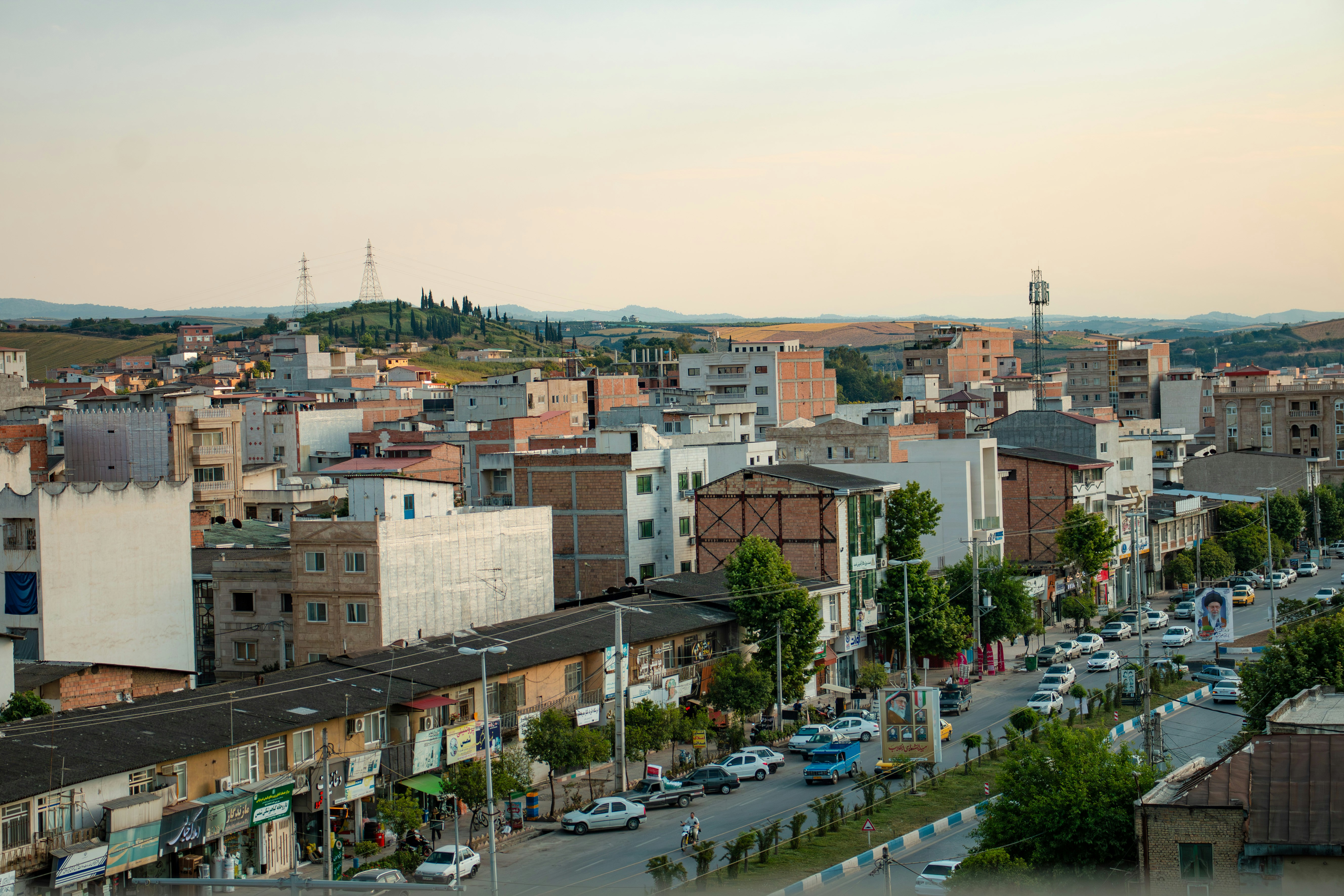 A mid-rise urban street scene viewed from a gentle aerial angle, with buildings, trees, and moving traffic under a clear sky.