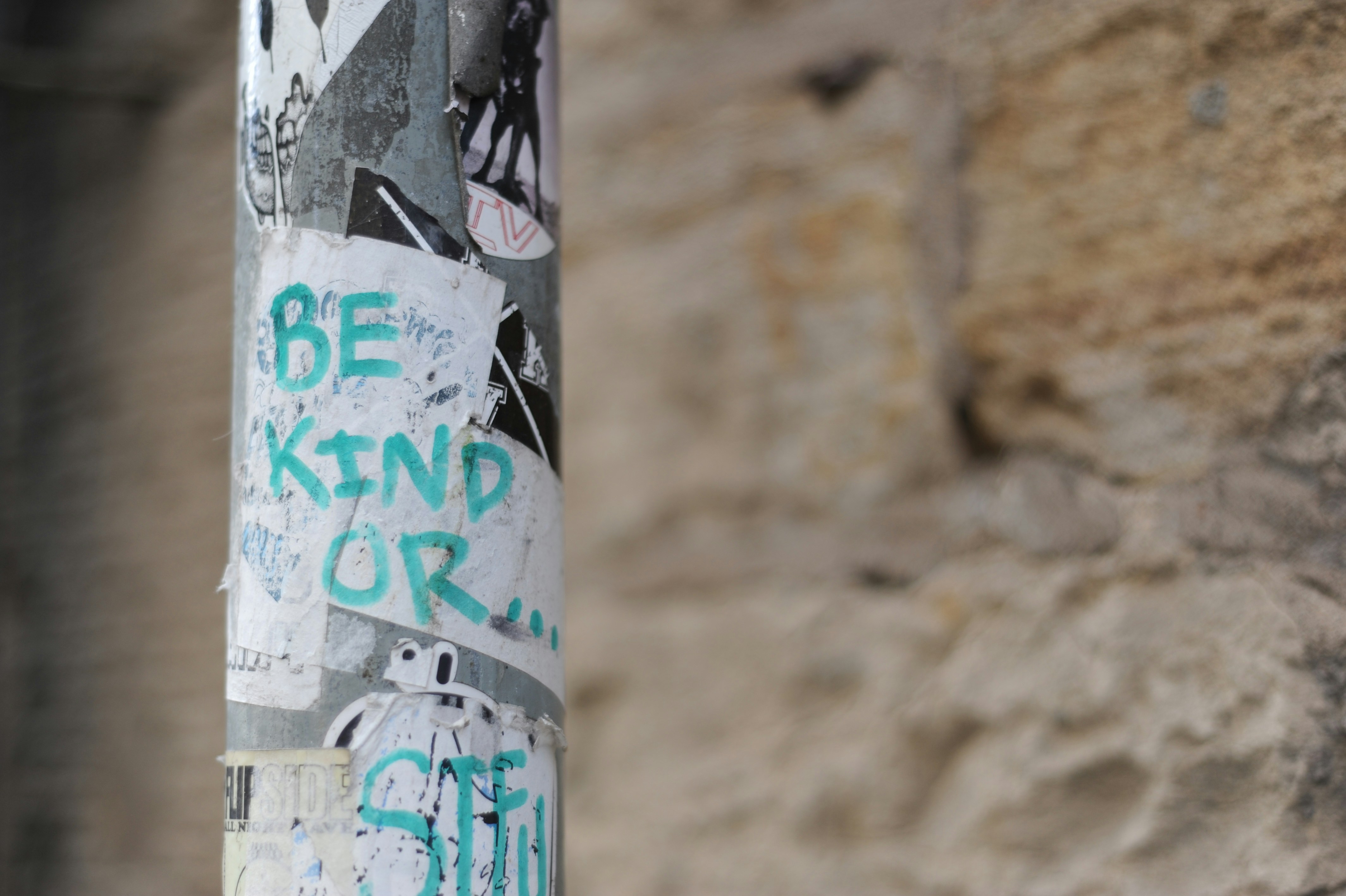 A grey metal lamppost wrapped in layered stickers, with hand-lettered "BE KIND OR..." dominating the frame. Below, a "STFU" scrawl is partly visible. Out-of-focus stone wall behind gives the post the feel of a small urban monument.