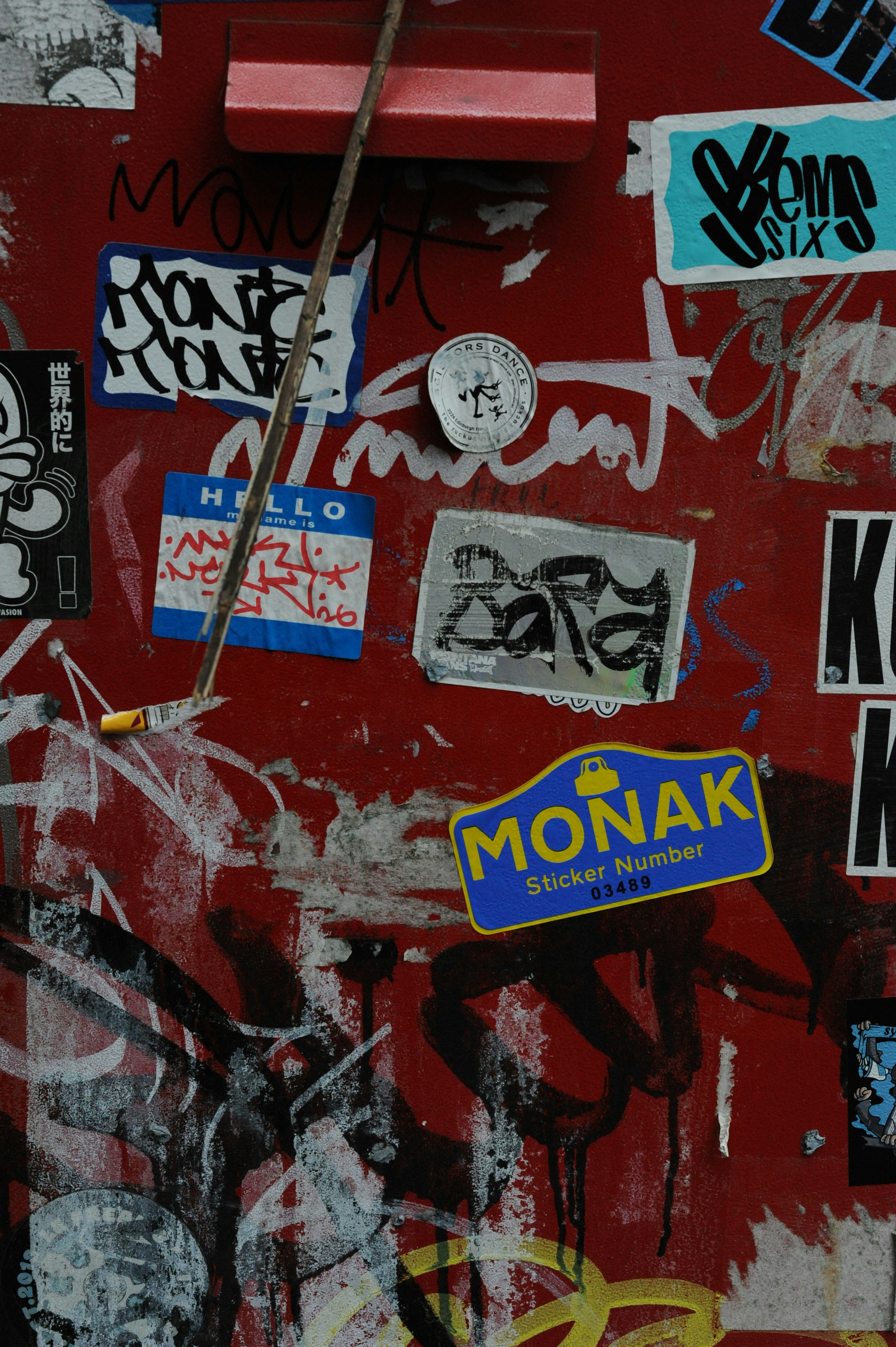A close-up of a red telephone or utility box plastered in layered street stickers, including a yellow "MONAK Sticker Number 03489" tag, a Hello My Name Is sticker, and a black scrawled tag reading "innocent". Drips of white paint streak across the surface like graffiti residue.