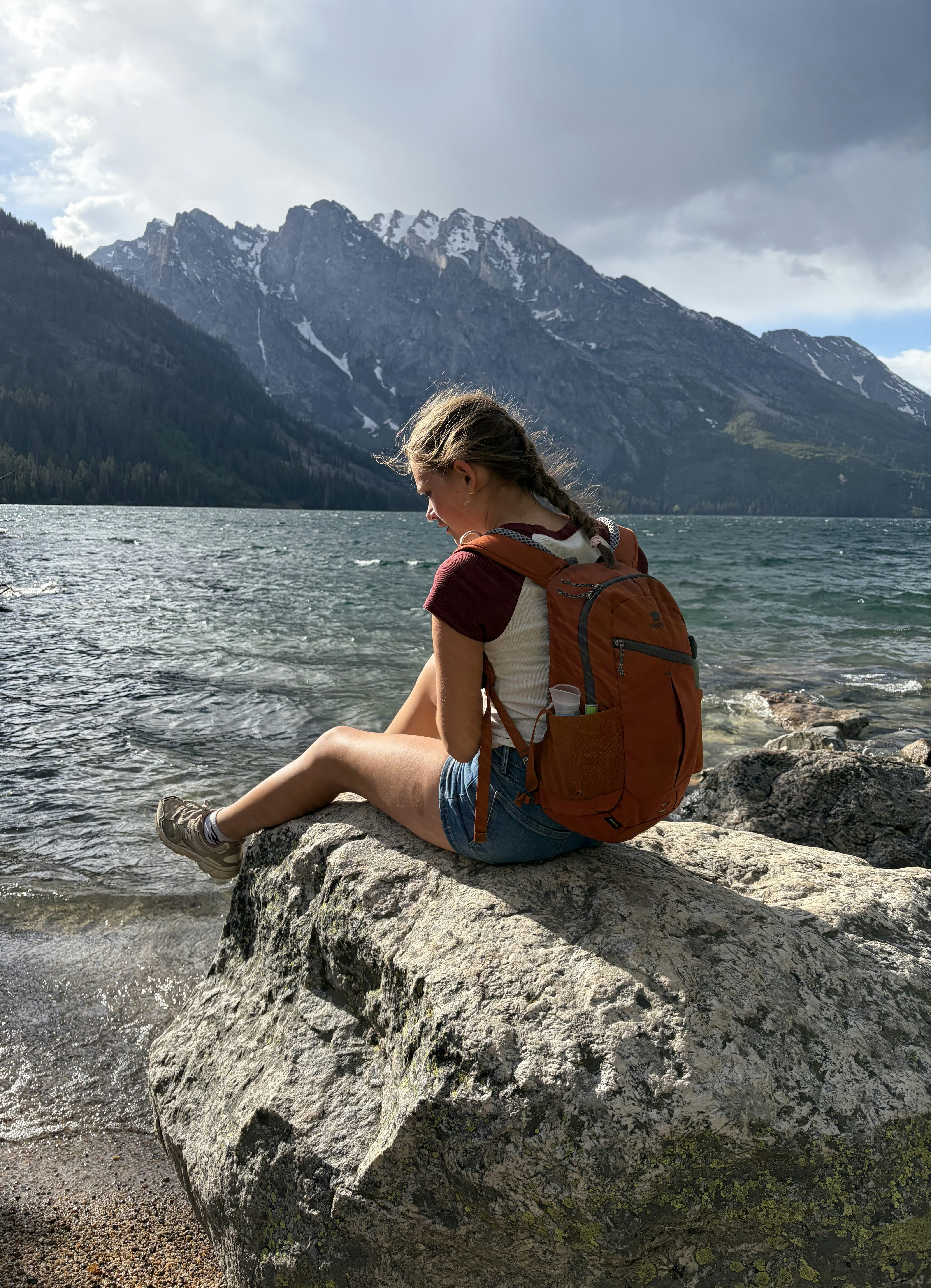 Young hiker resting on a rock by a lake with rugged mountains in the background; orange backpack stands out against the landscape. A photograph capturing a peaceful outdoor moment.