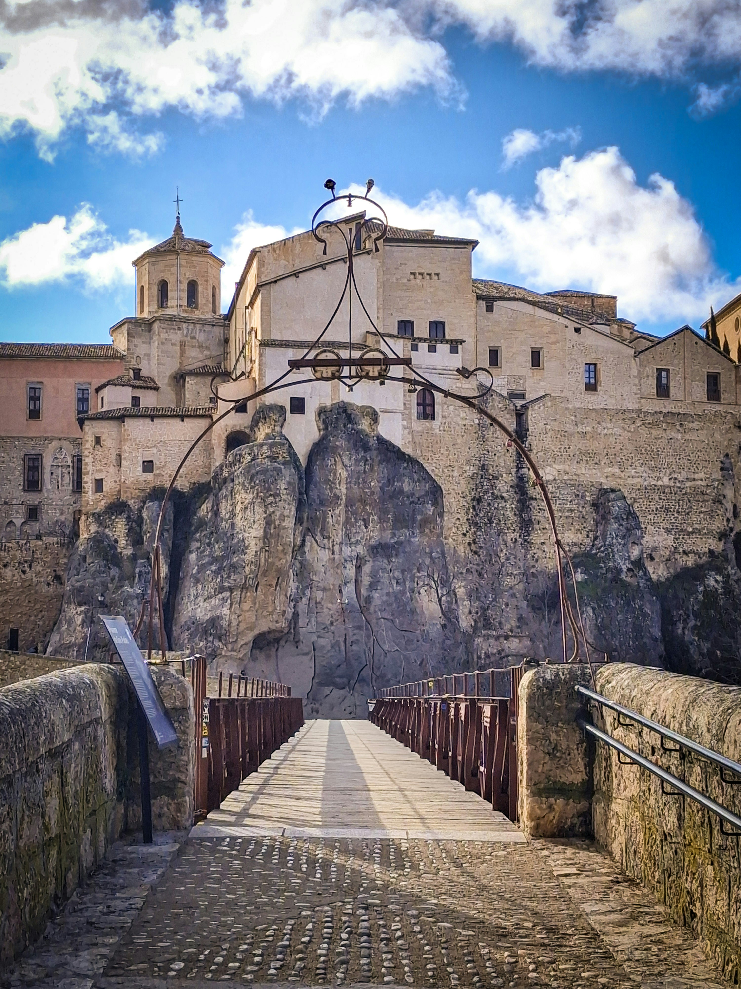 A low-angle perspective of the historic San Pablo iron footbridge leading toward the medieval cliffside town of Cuenca, Spain, under a blue sky with white clouds.