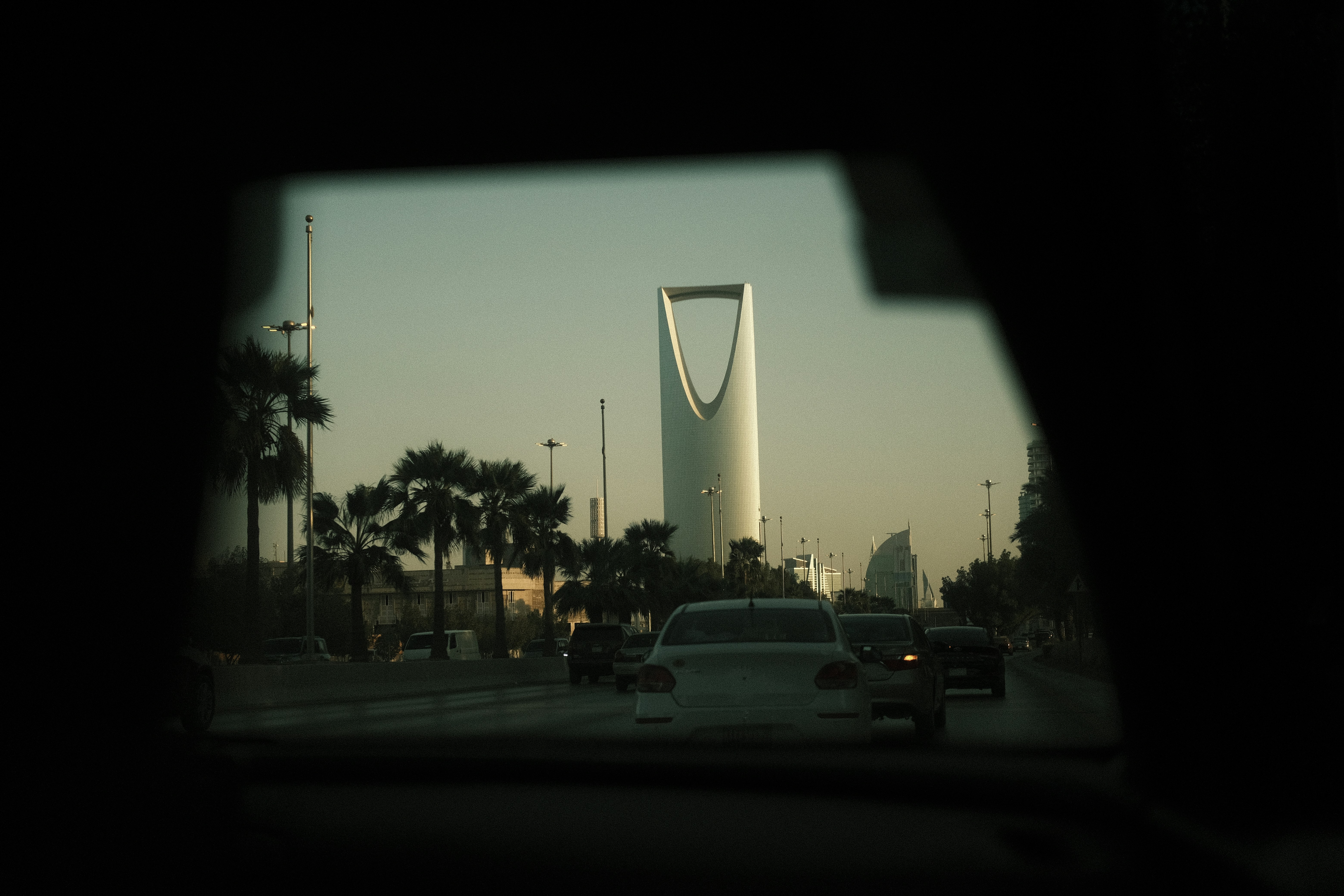 Modern skyscraper dominates city skyline with palm trees and cars.