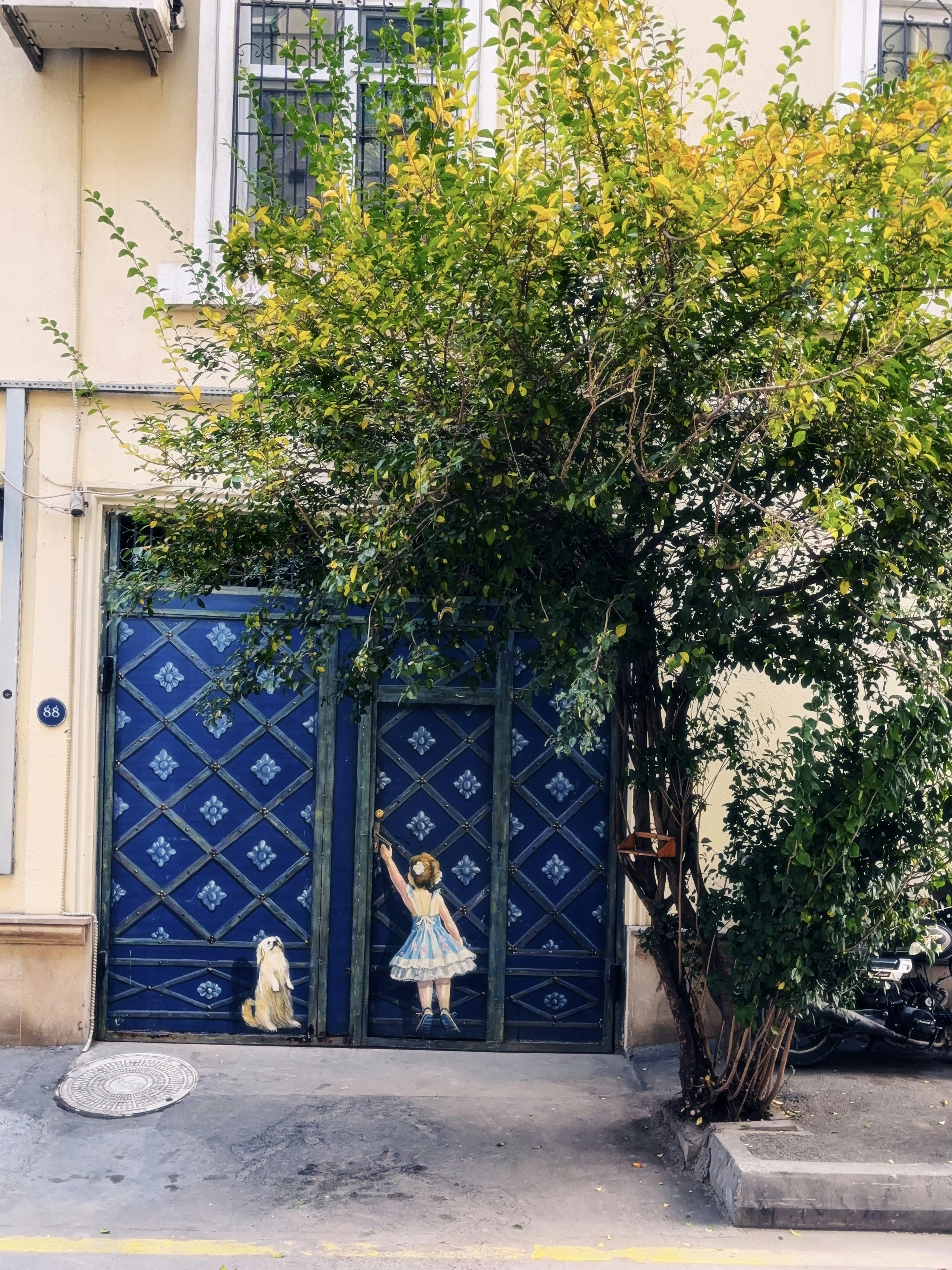A girl in a blue dress stands by a blue patterned gate, accompanied by a light-colored dog, with a lush tree arching over the entrance in an urban setting.