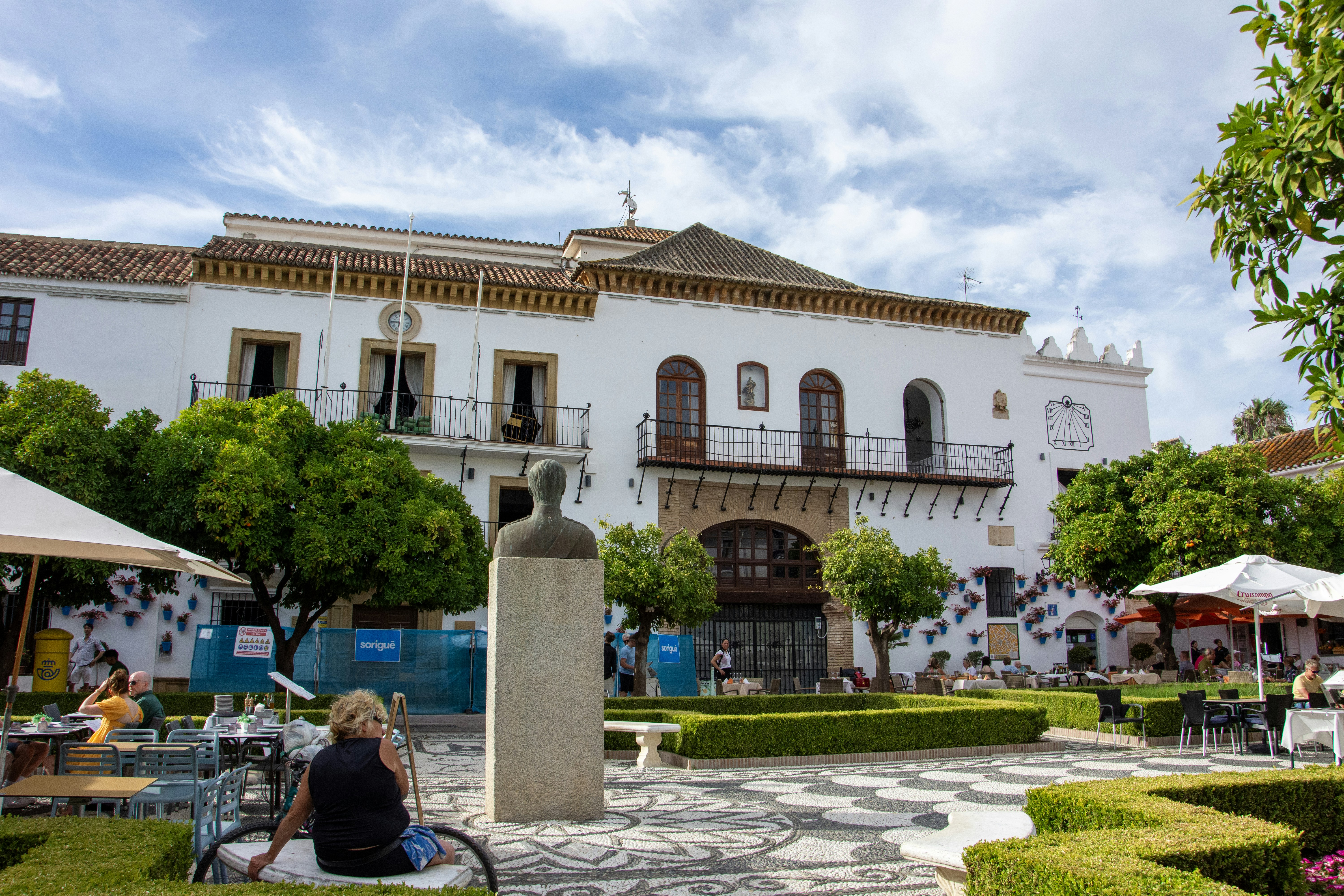 A sunlit plaza in front of a white colonial building with a statue centerpiece
