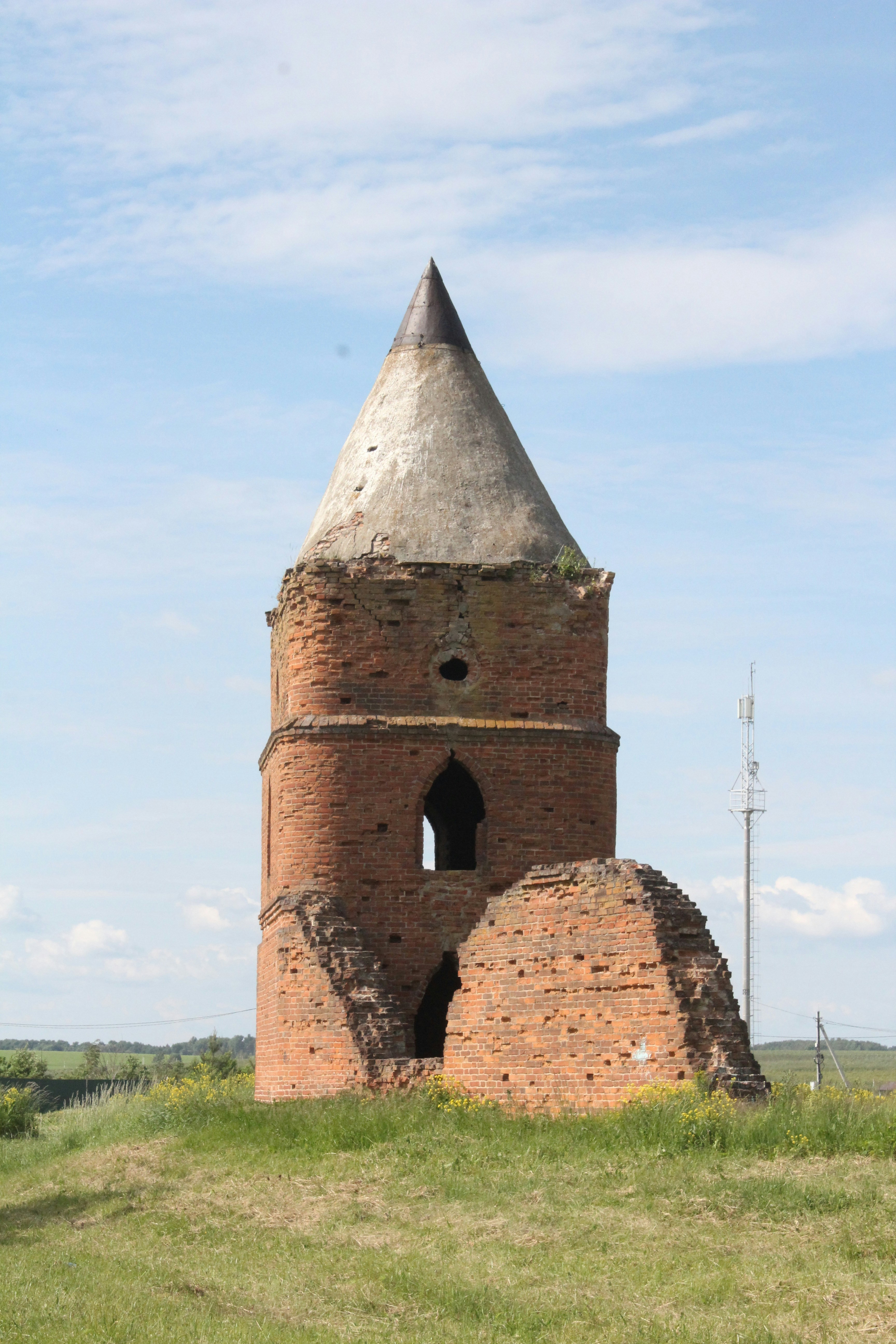 Ruined brick tower with conical roof standing in an open field under a blue sky