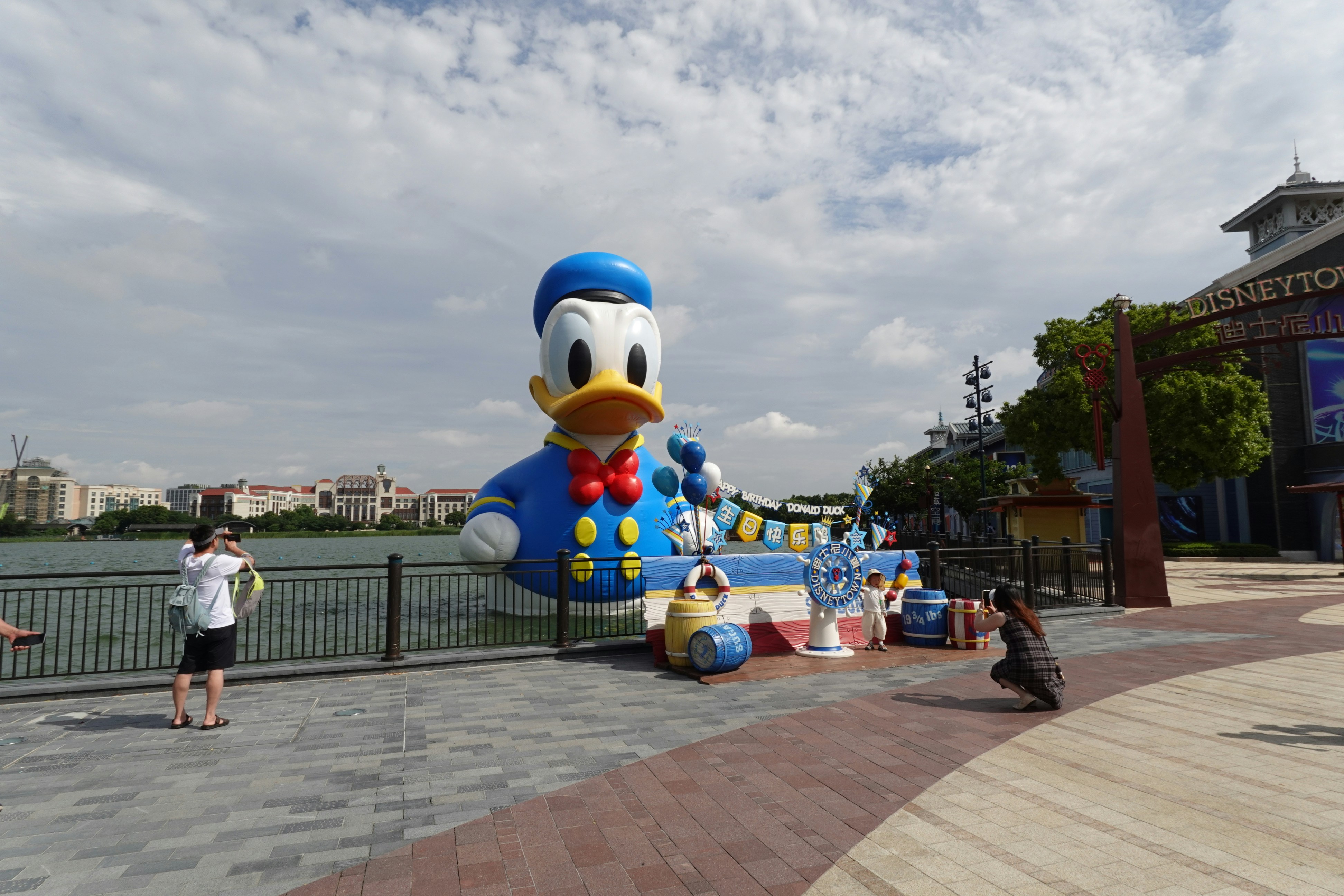 Colorful Donald Duck statue with surrounding decorative props along a stone-paved waterfront promenade near a Disney-themed venue.