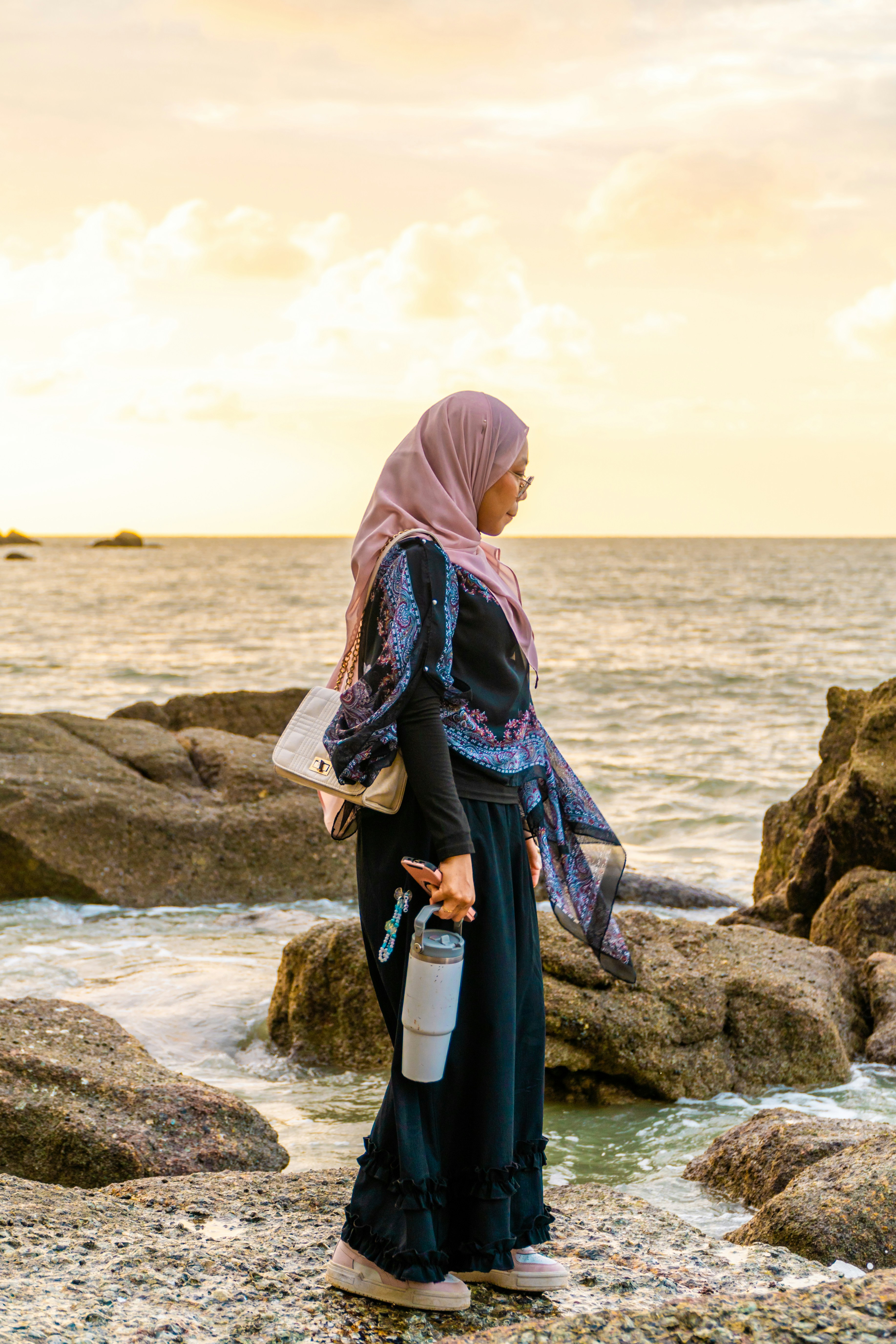 Woman in hijab standing on rocky beach at sunset.