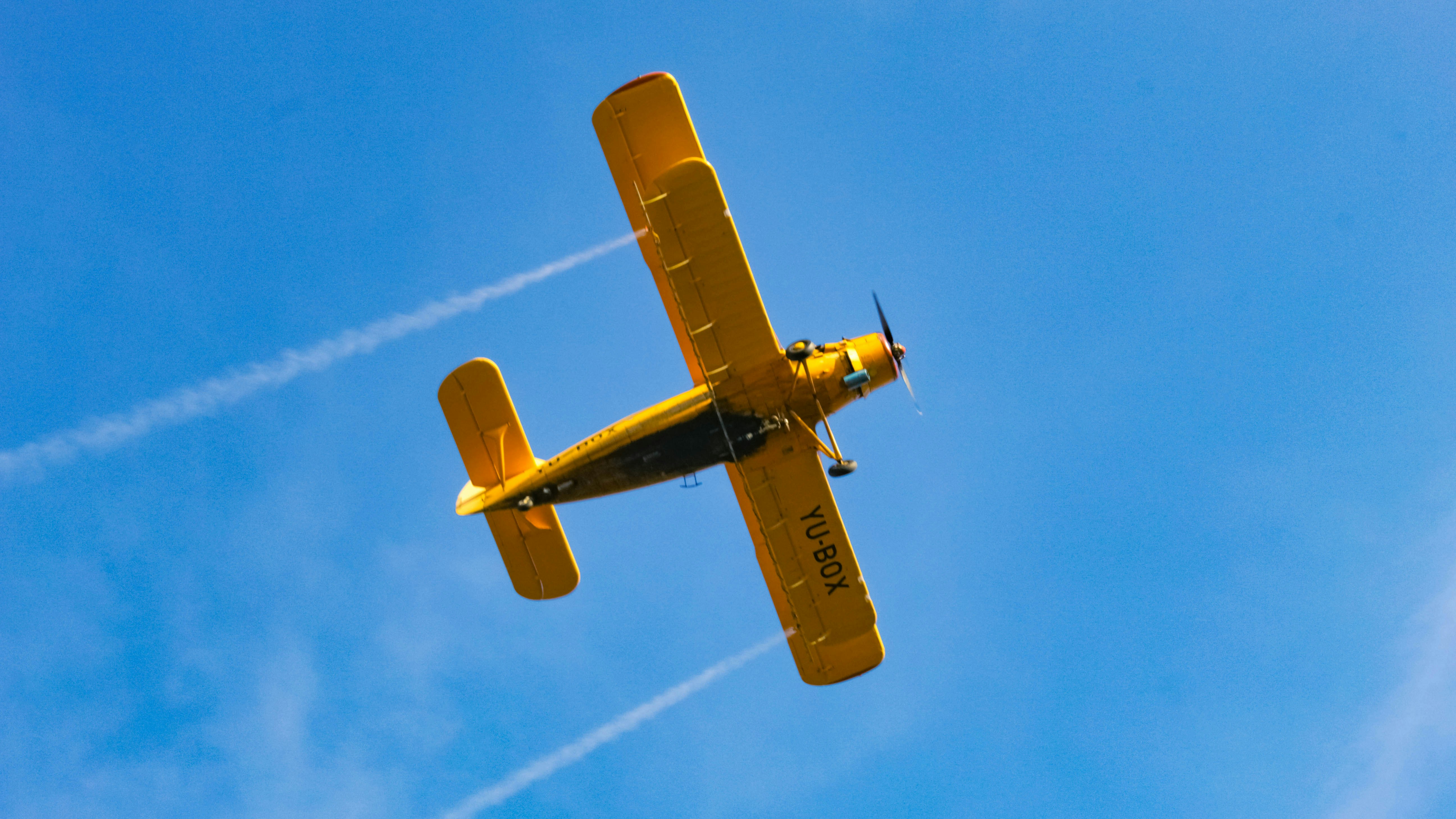 A yellow biplane flying in a blue sky, leaving contrails.