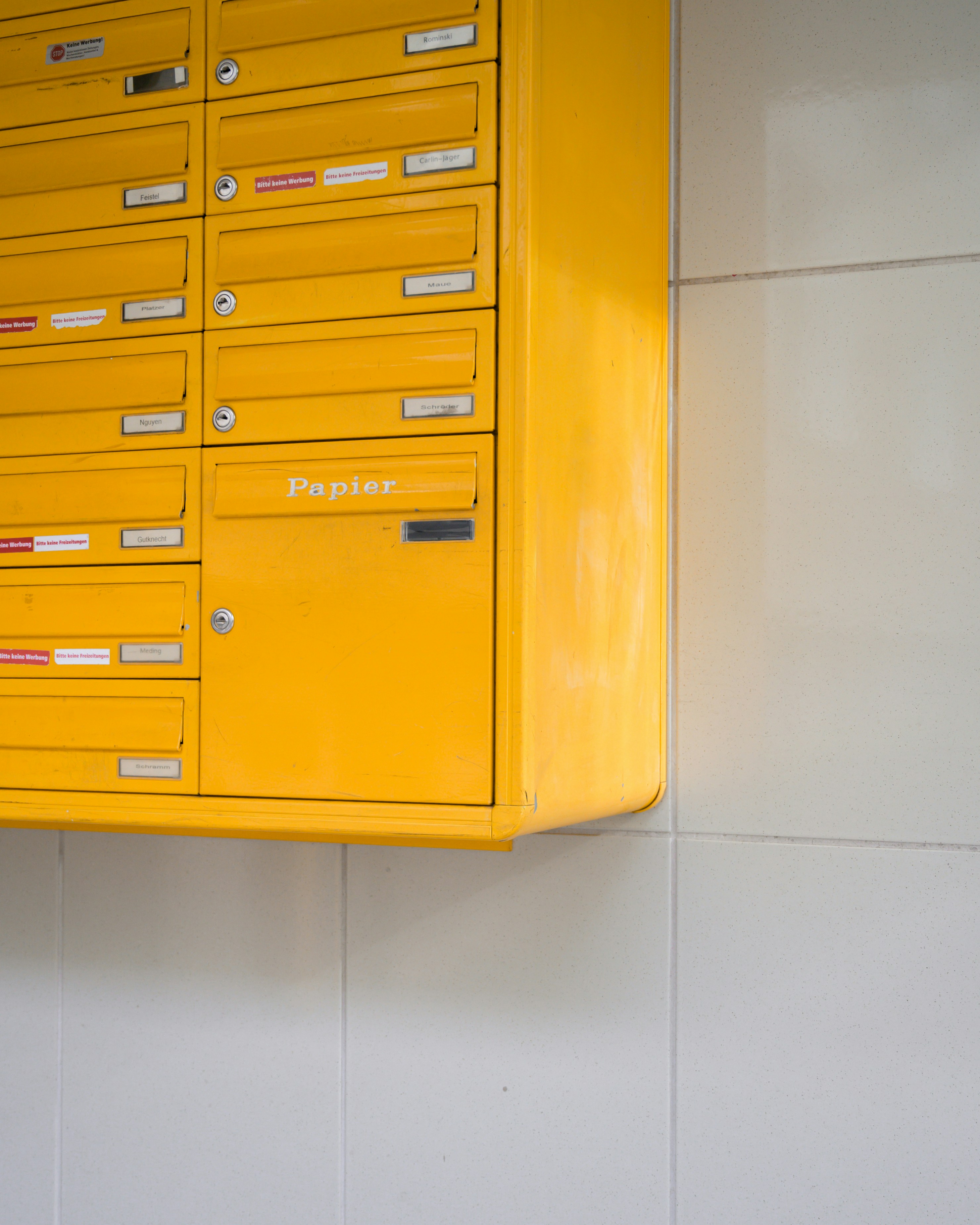 Bright yellow mailbox cluster mounted on a pale tiled wall, with a single shadowed edge and a small label; emphasizes color and urban utility without readable text.