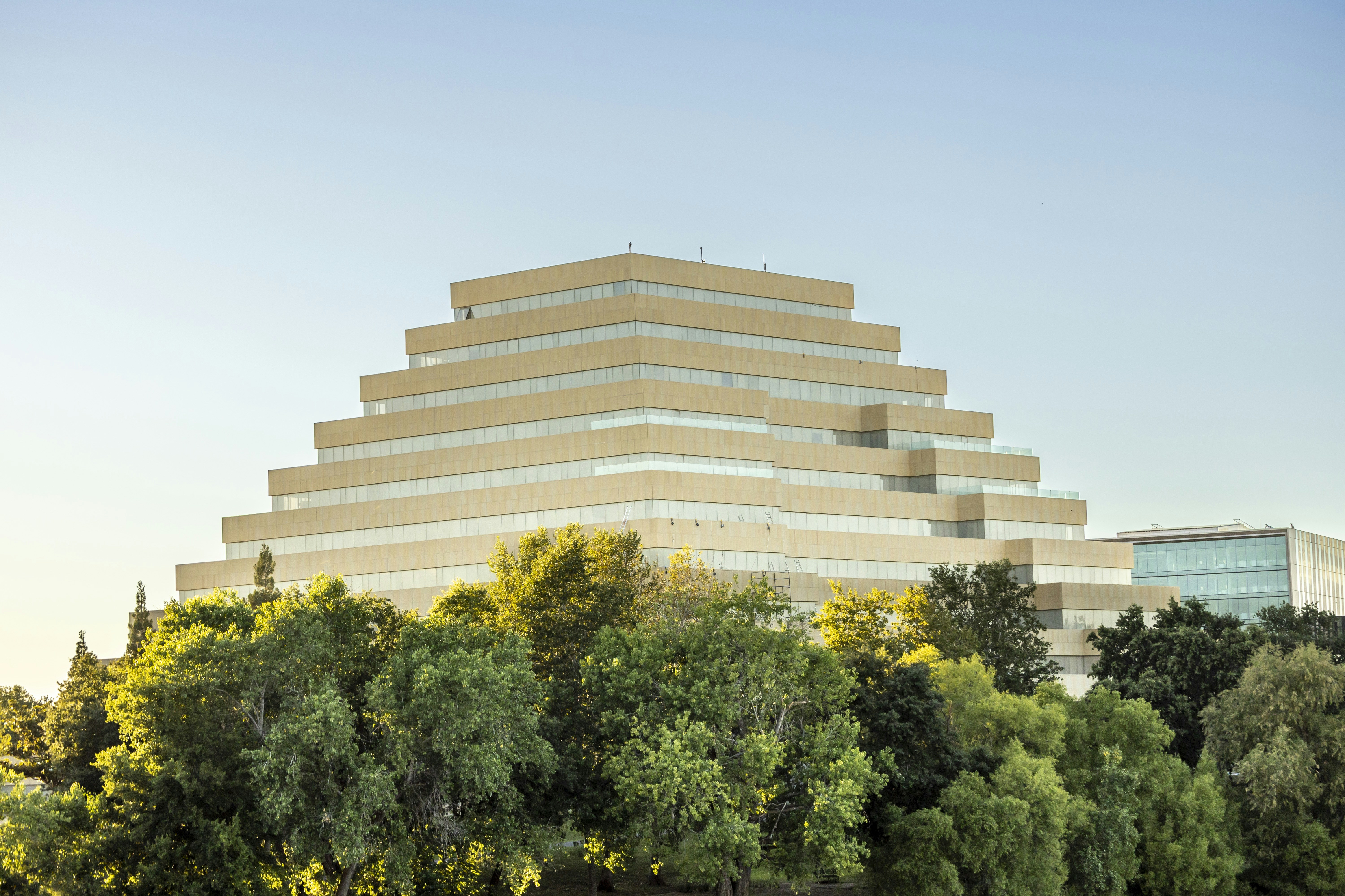 Terraced, pyramid-like building rises above a lush tree line under a clear sky, showcasing geometric architecture. This image captures the structure’s layered, stepped facade with surrounding greenery.