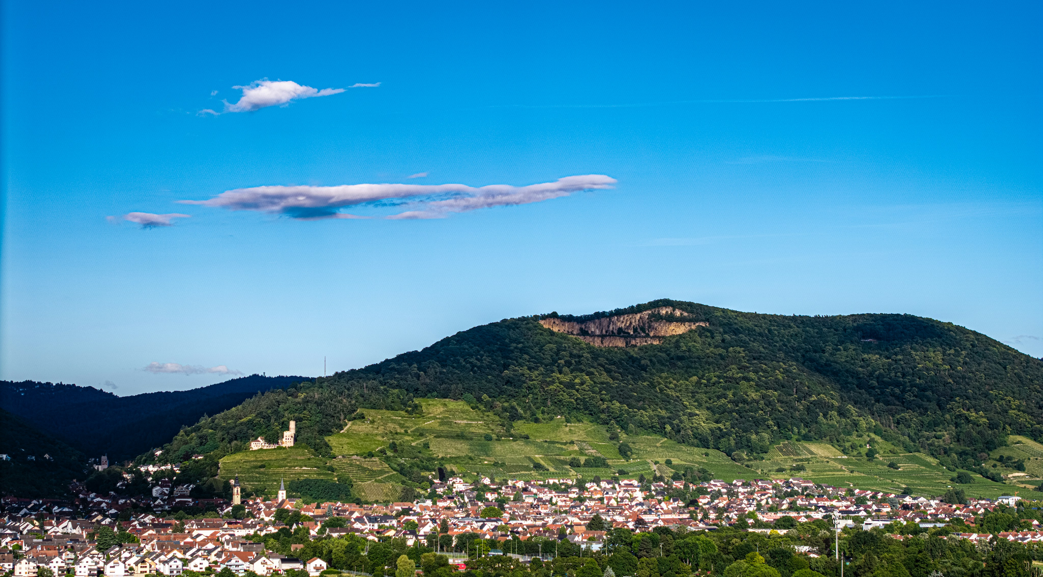 Town nestled below a large green mountain under blue sky.