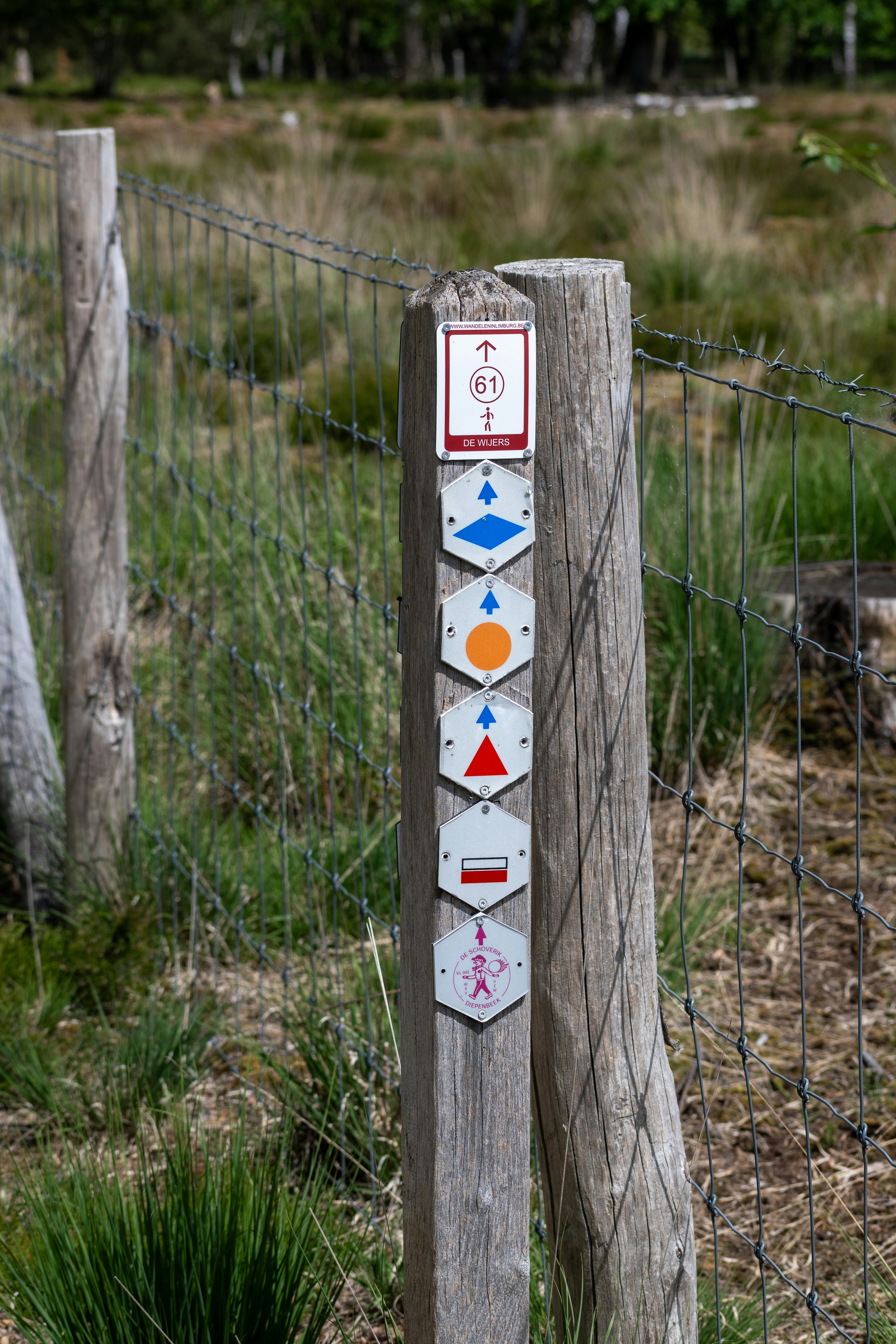 Marker signs climbing a wooden post beside a wire fence in a grassy field with a distant tree line.