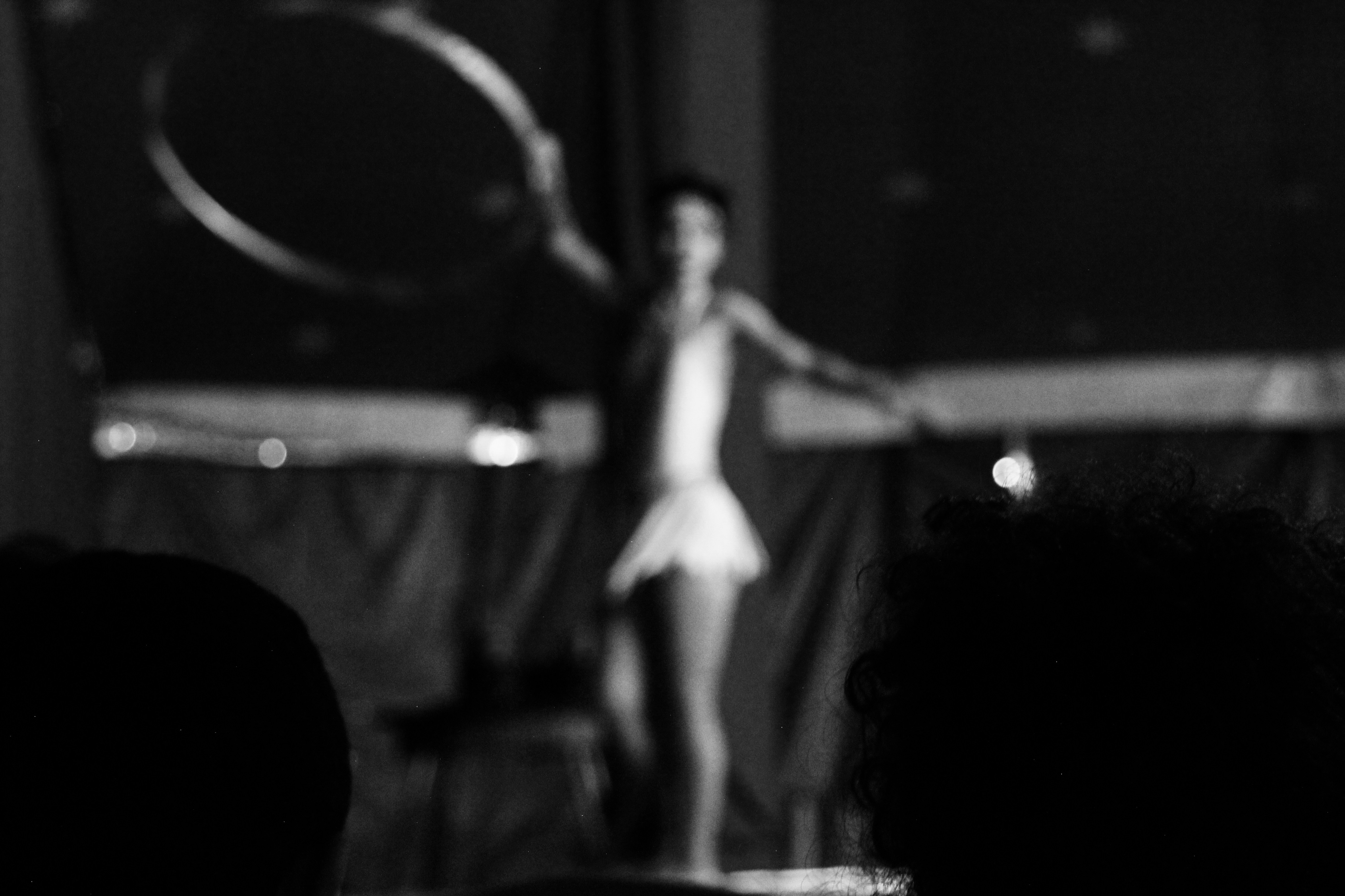 Stage performer in a blurred, monochrome scene with an out-of-focus audience silhouette; a circular prop and draped background suggest a rehearsal or performance setting.