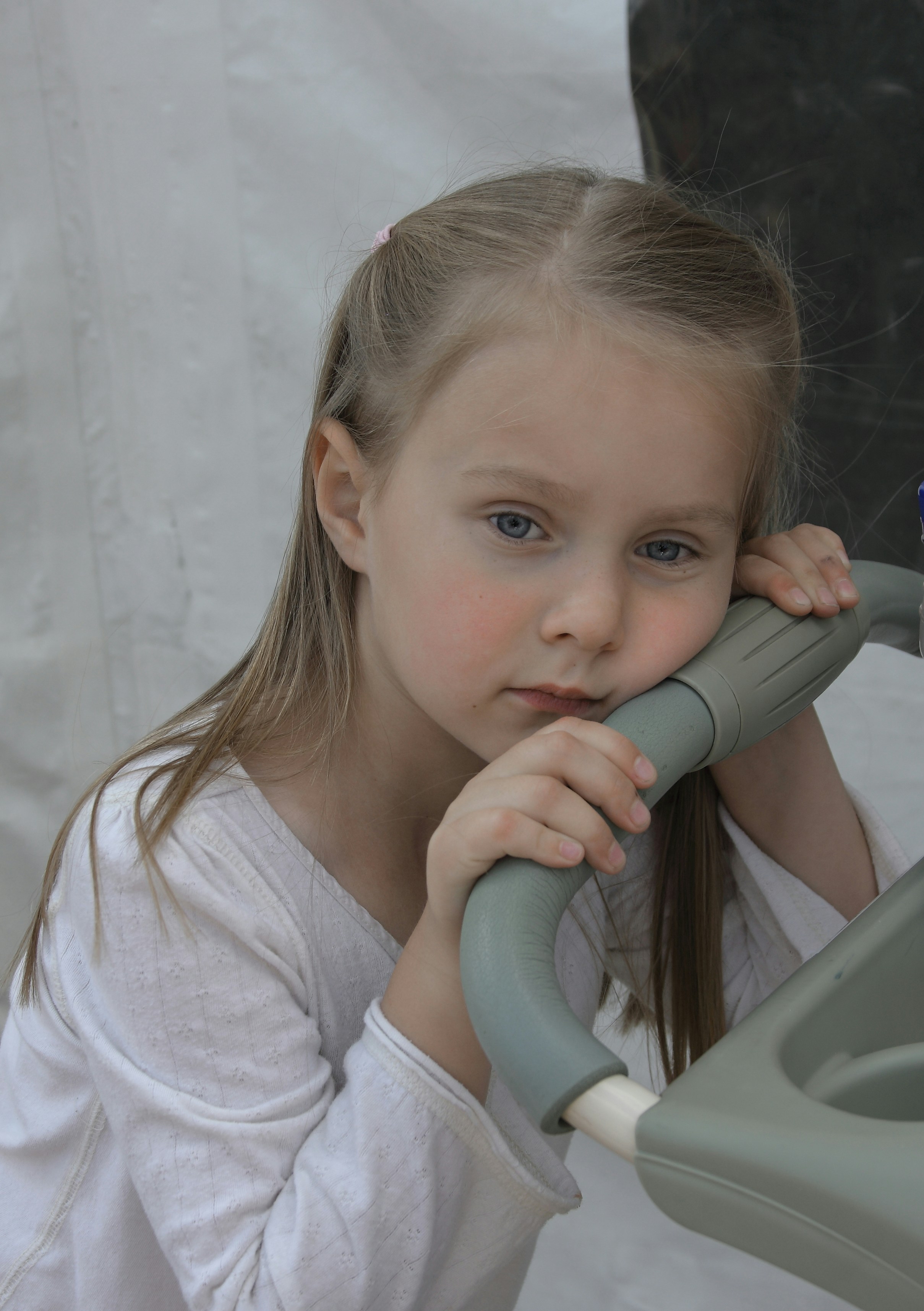 A young girl leans against a stroller bar, gazing softly at the camera with a calm expression; the scene is a close, gentle studio-style portrait with natural lighting.