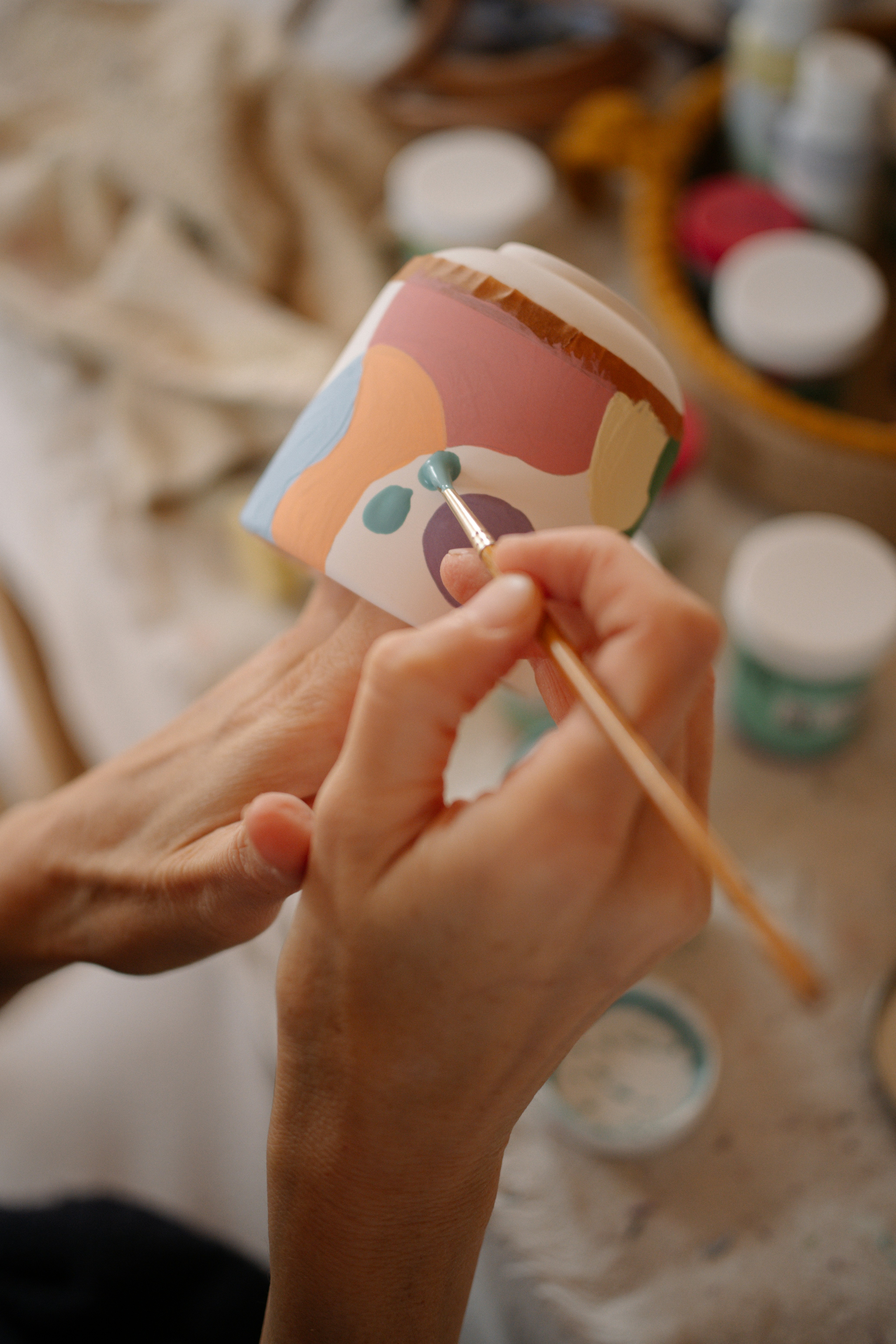 A person's hands painting colorful designs on a ceramic cup.