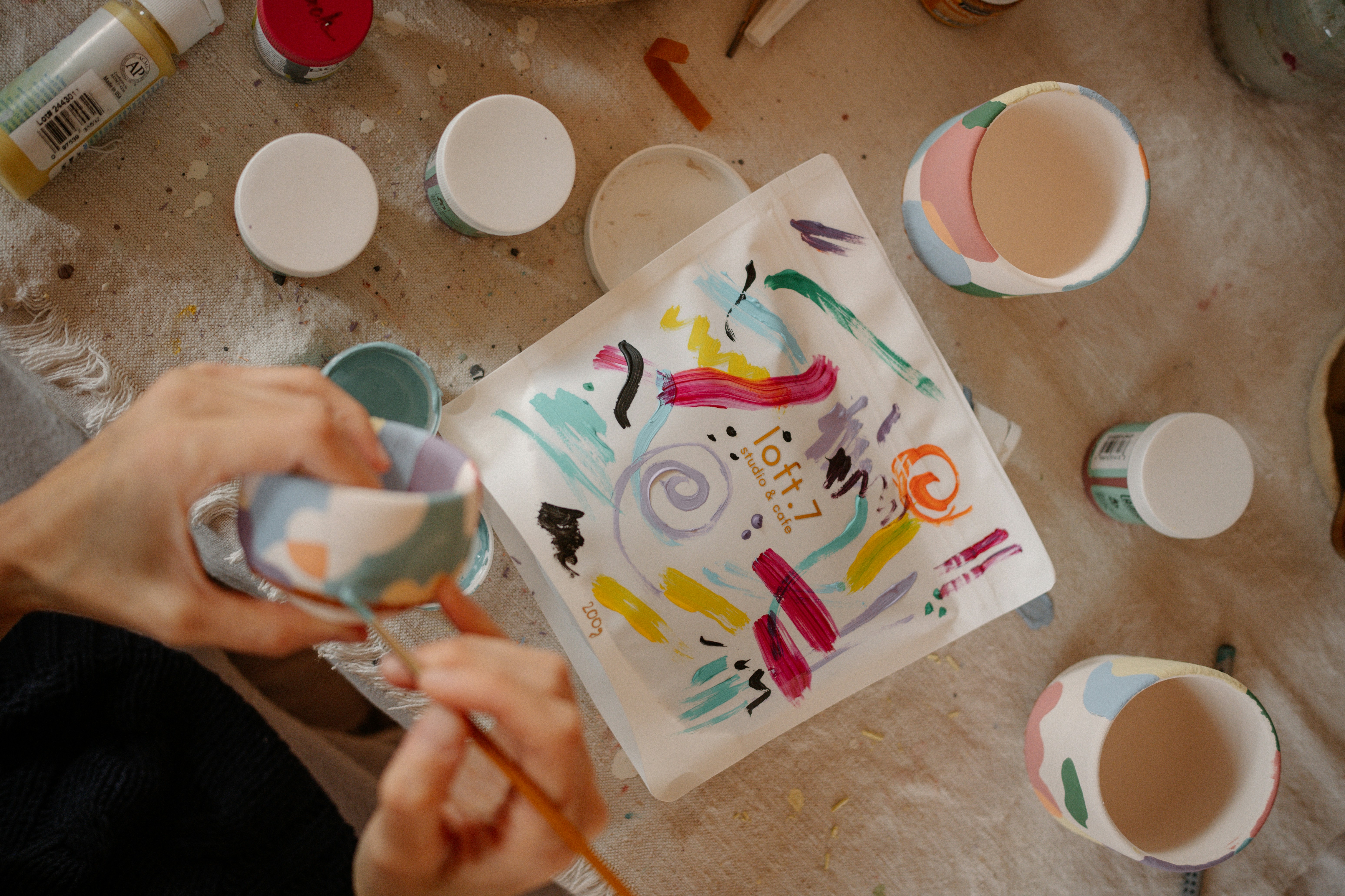 Hands painting colorful abstract designs on ceramic cups.