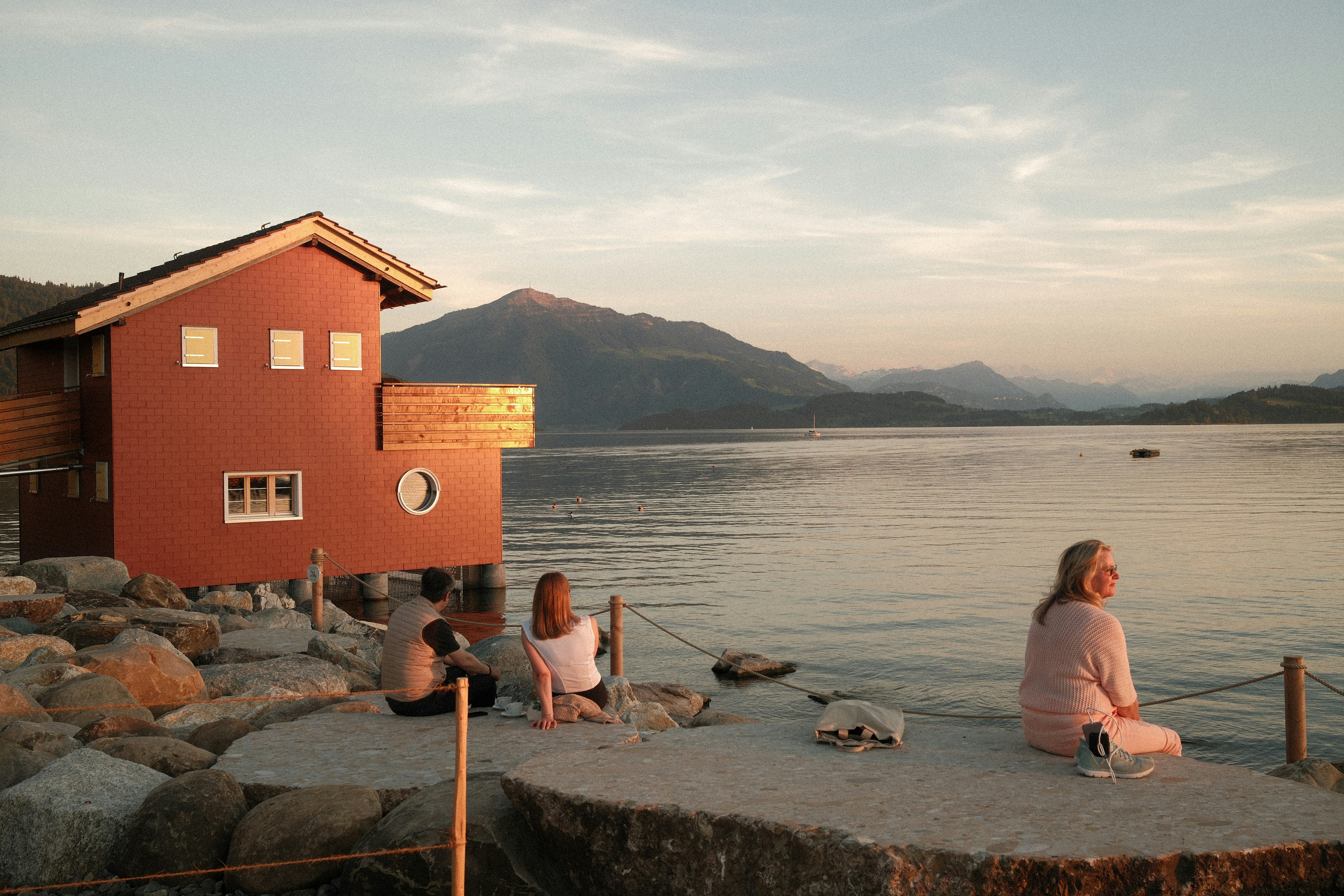 People relaxing by a red house and lake at sunset.