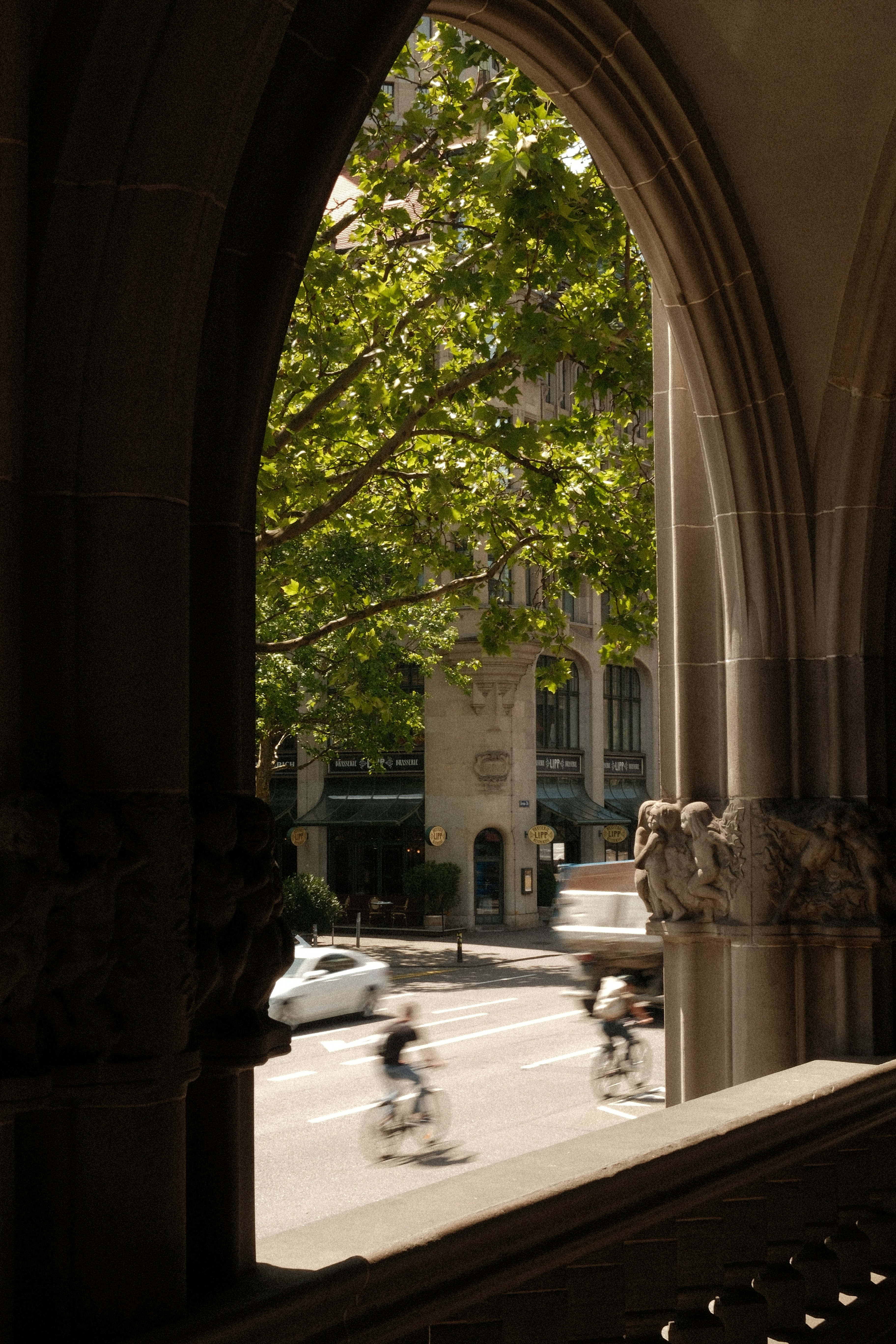 City street with cyclists and car viewed through stone archway.