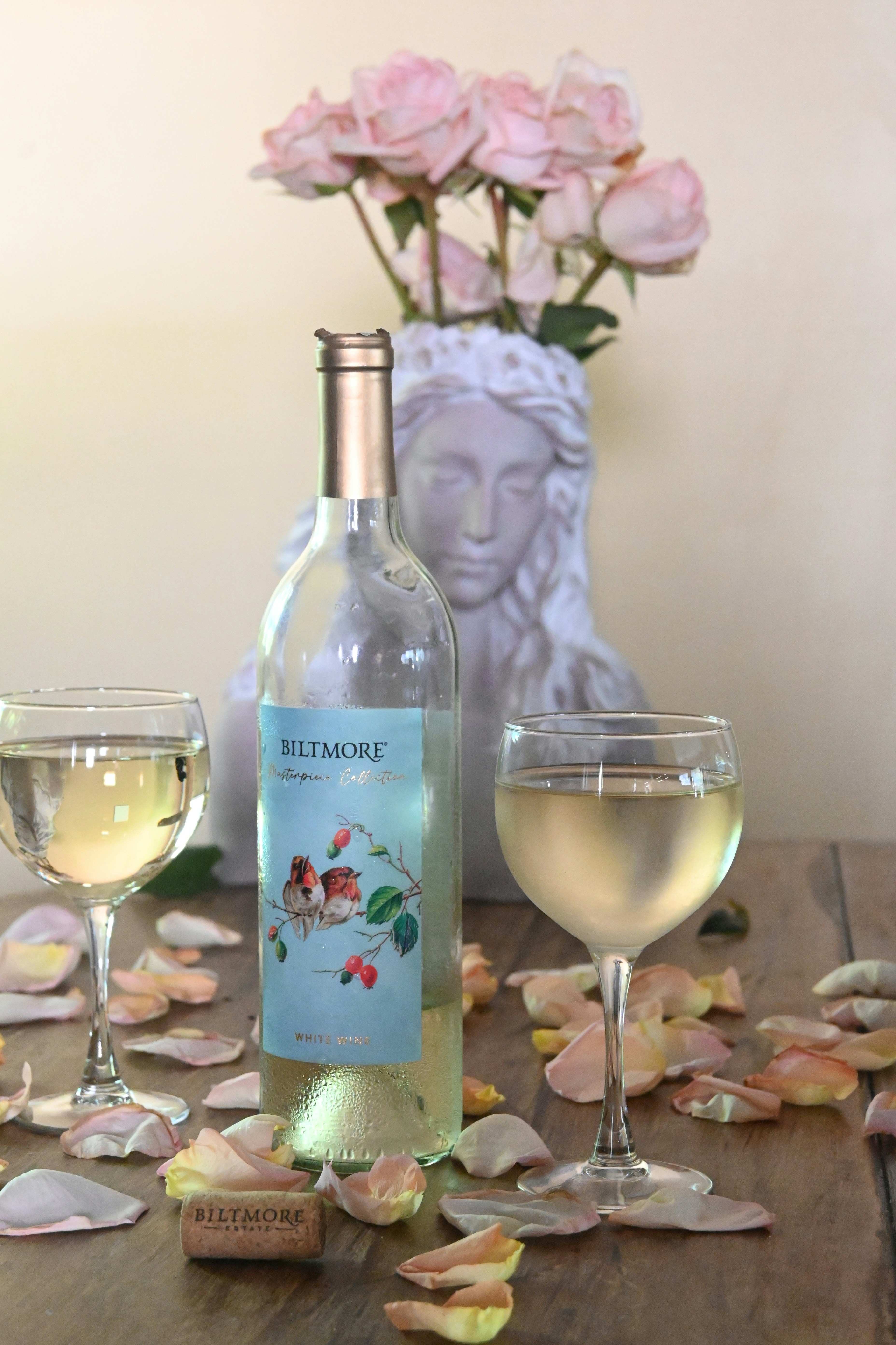 White wine bottle, two glasses, rose petals, and a statue.