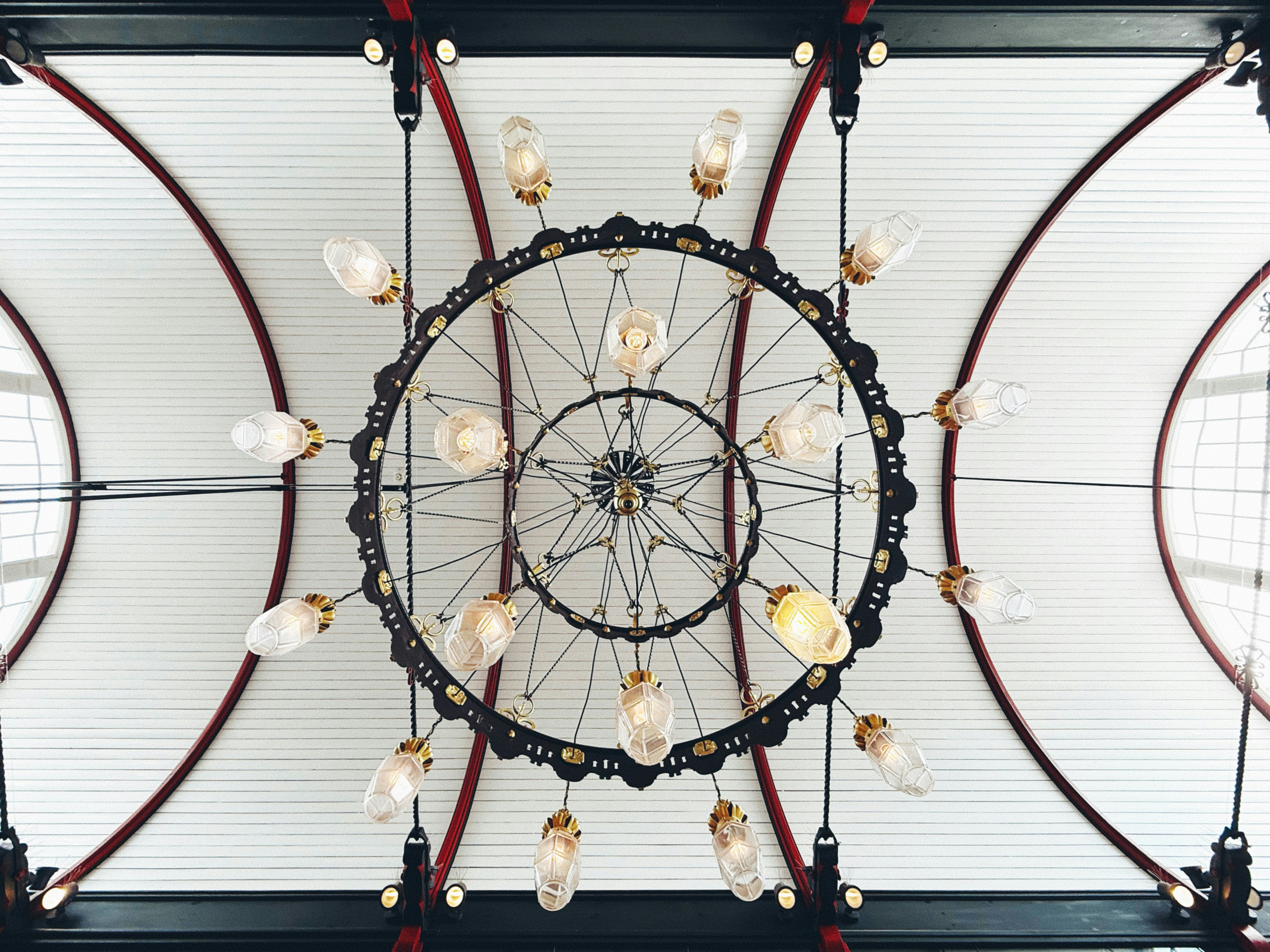 The photo captures an intricate chandelier featuring a circular design with multiple light fixtures resembling bicycle wheels, hanging from a ceiling with red arches and white paneling.