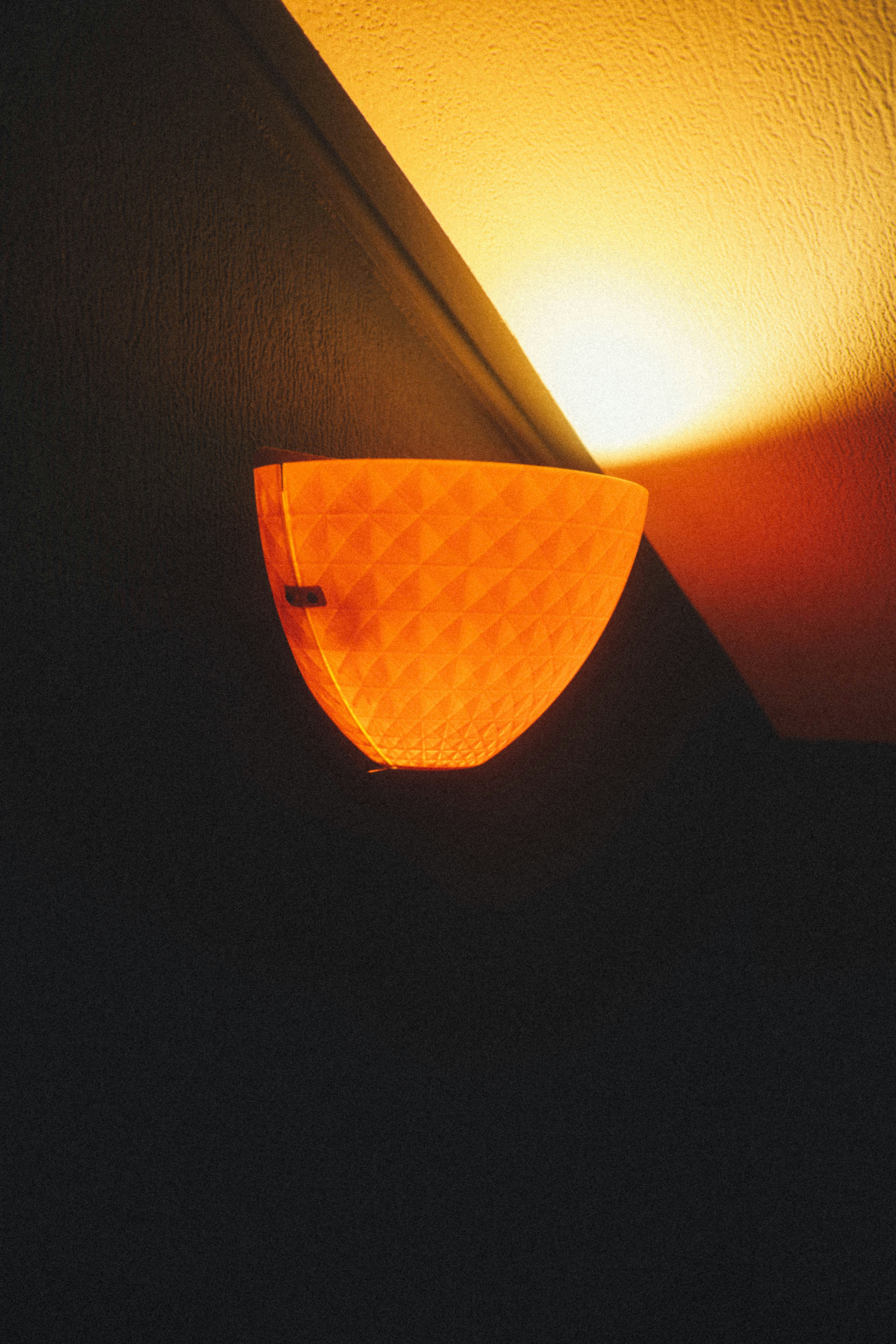 A textured amber lampshade casts a soft glow on a dark wall, creating a simple interior vignette with strong contrast and geometric pattern. The scene emphasizes warm light and minimal composition.