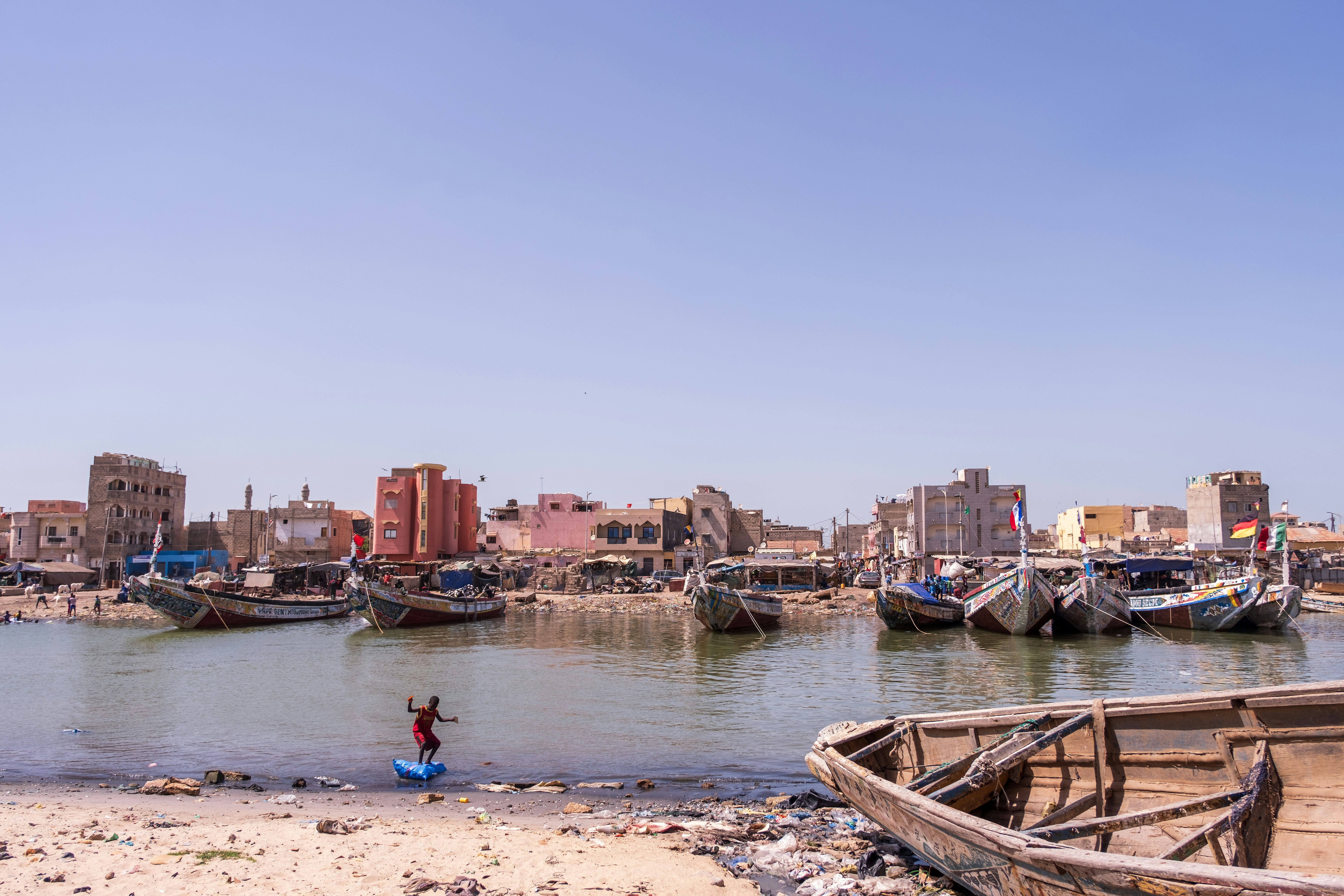This wide-angle photograph captures a vibrant waterfront scene in Saint-Louis, Senegal, under a clear, expansive sky. In the foreground, an empty, weathered wooden hull rests on the sandy bank, while across the river, a large fleet of traditional painted pirogues is moored along the opposite shore. The background showcases the colorful, densely packed buildings of the coastal town, and a local child playfully balances on a blue block in the shallow water, adding life to the scene.